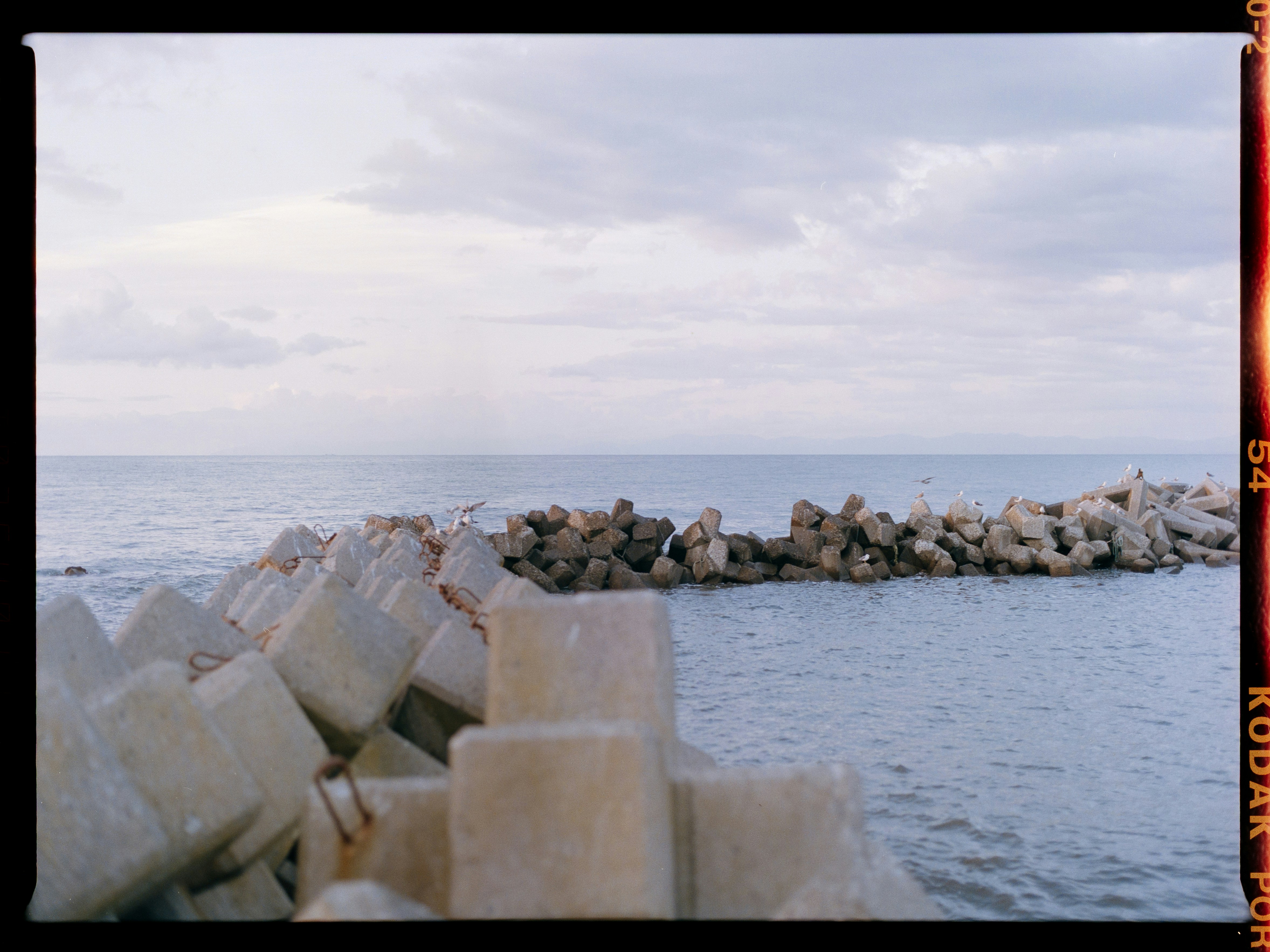 Concrete breakwaters jutting into the calm sea under a soft, cloudy sky.