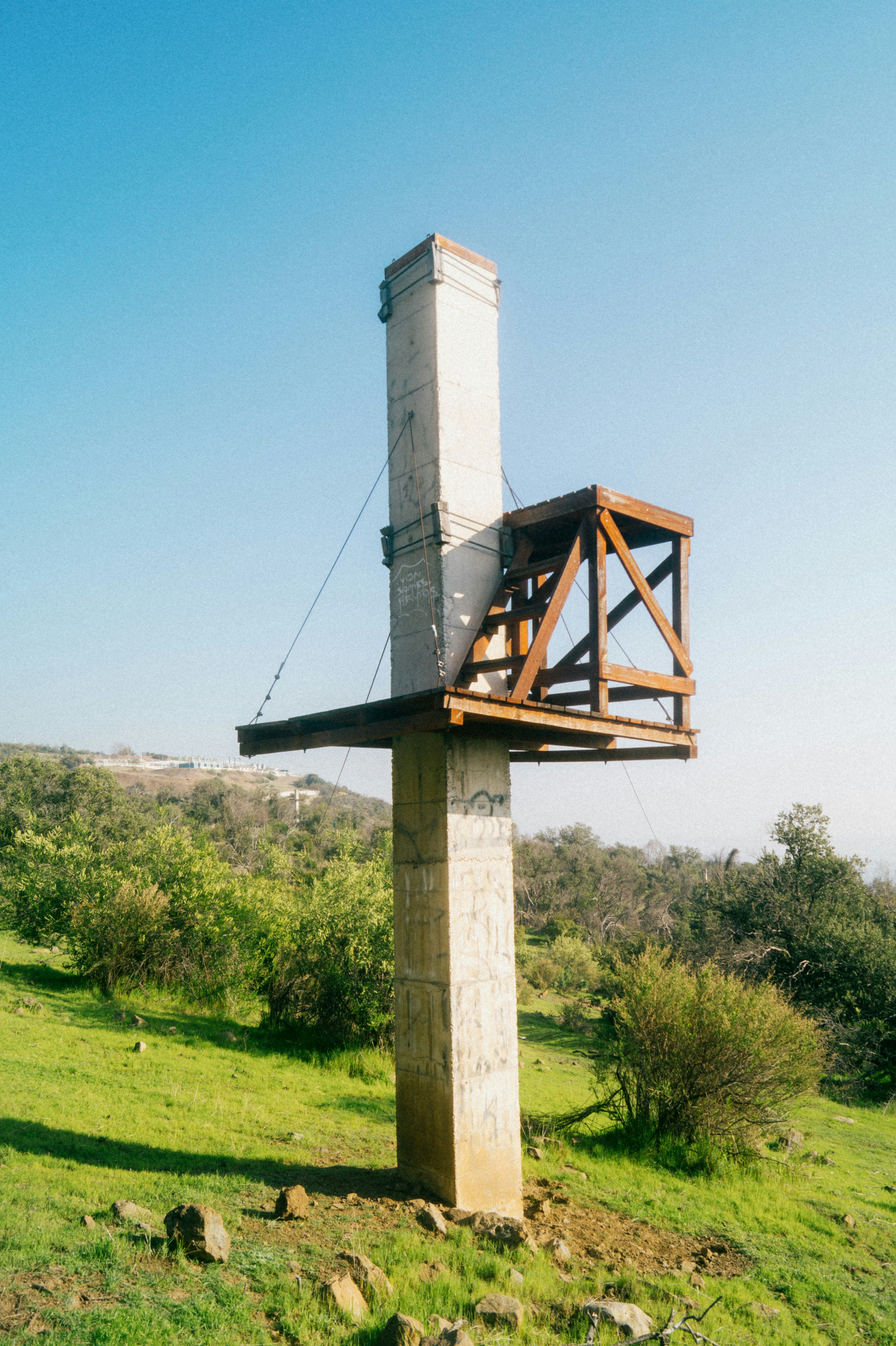 A solitary concrete structure with a wooden platform stands amidst lush greenery under a clear blue sky. This intriguing installation contrasts industrial elements with natural surroundings.