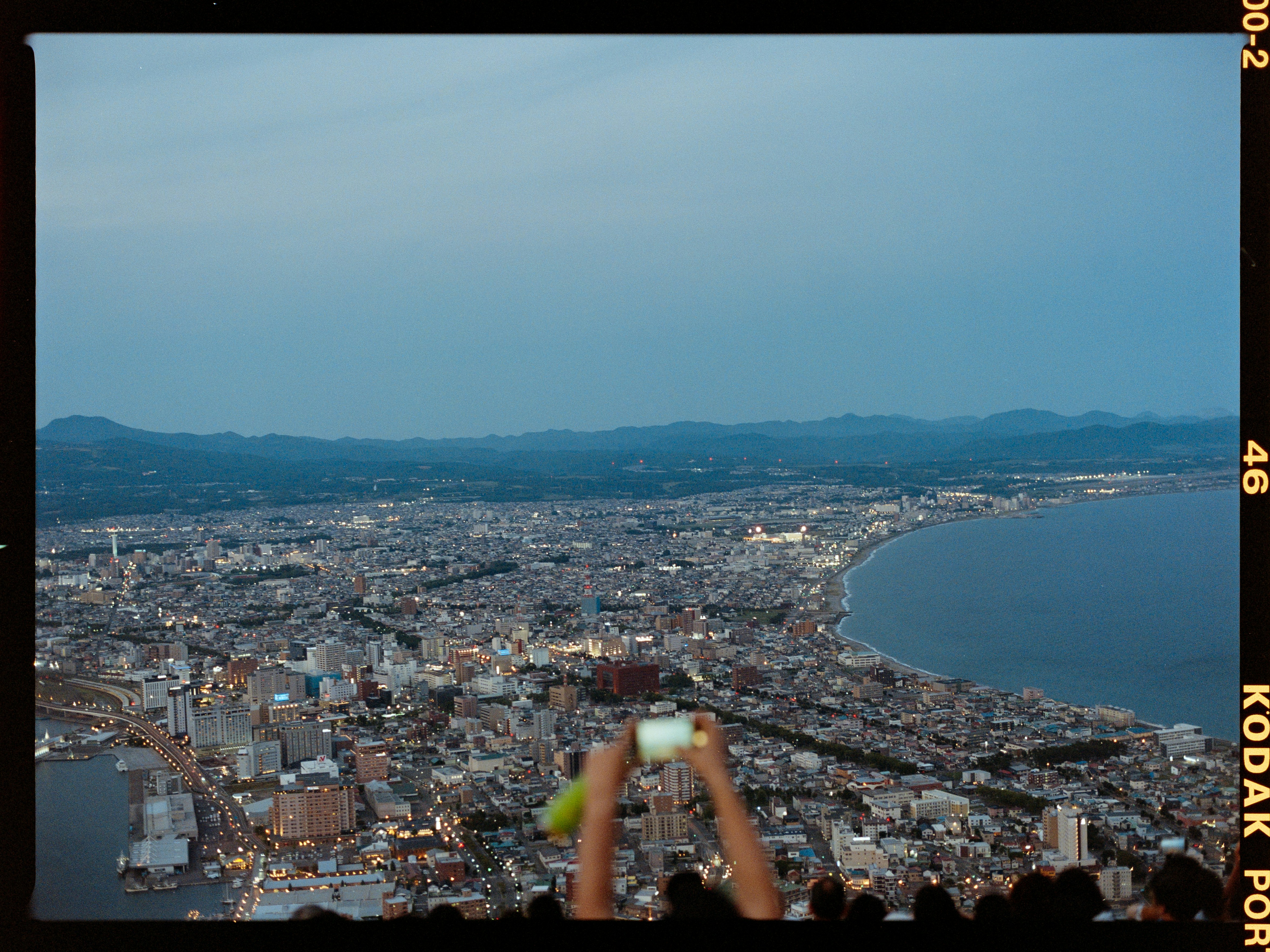 🎞️ Kodak Professional Portra 800｜🧪 Fuji Hunt C-41｜🖨️ EOS R6 + RF 100mm f/2.8L Macro｜📷 Mamiya 645 Pro + Mamiya-Sekor 80mm 1:1.9 | Person photographing a city skyline from above at dusk.