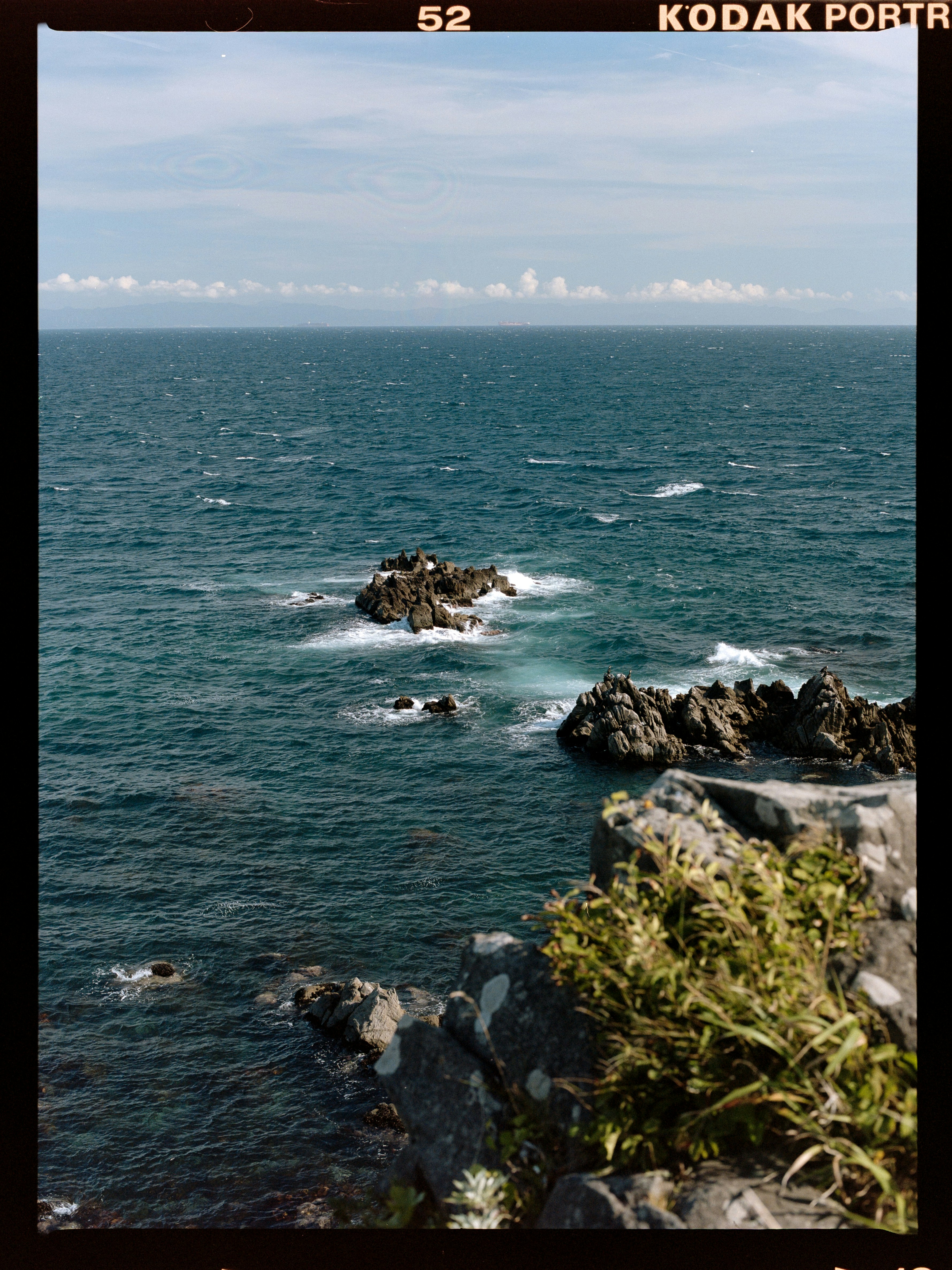 Rugged coastline with rocks emerging from vibrant blue waters, framed by lush greenery in the foreground.