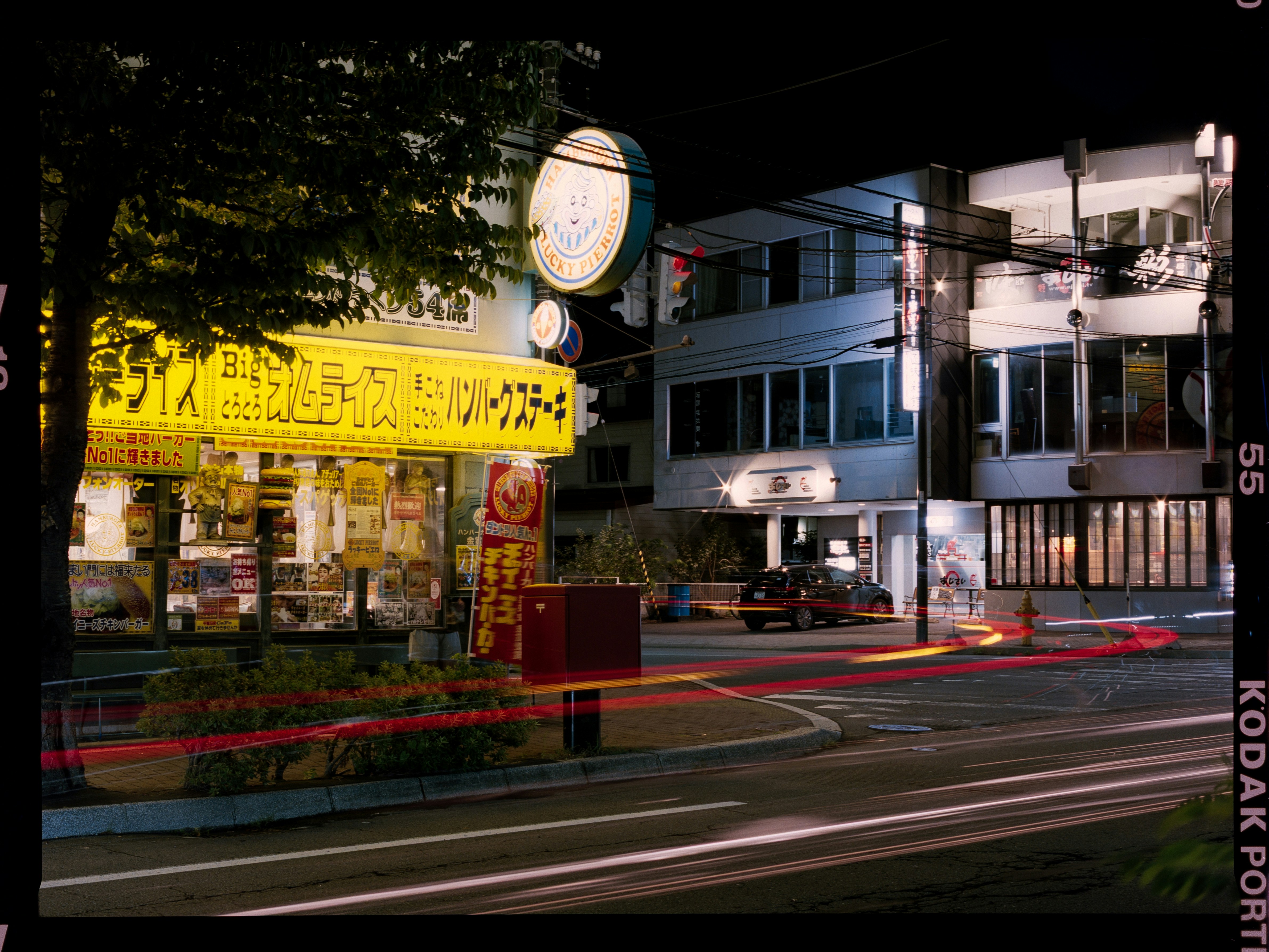 Brightly lit restaurant sign at night with vibrant yellow colors and passing car lights creating dynamic trails. The scene captures urban nightlife and dining options.