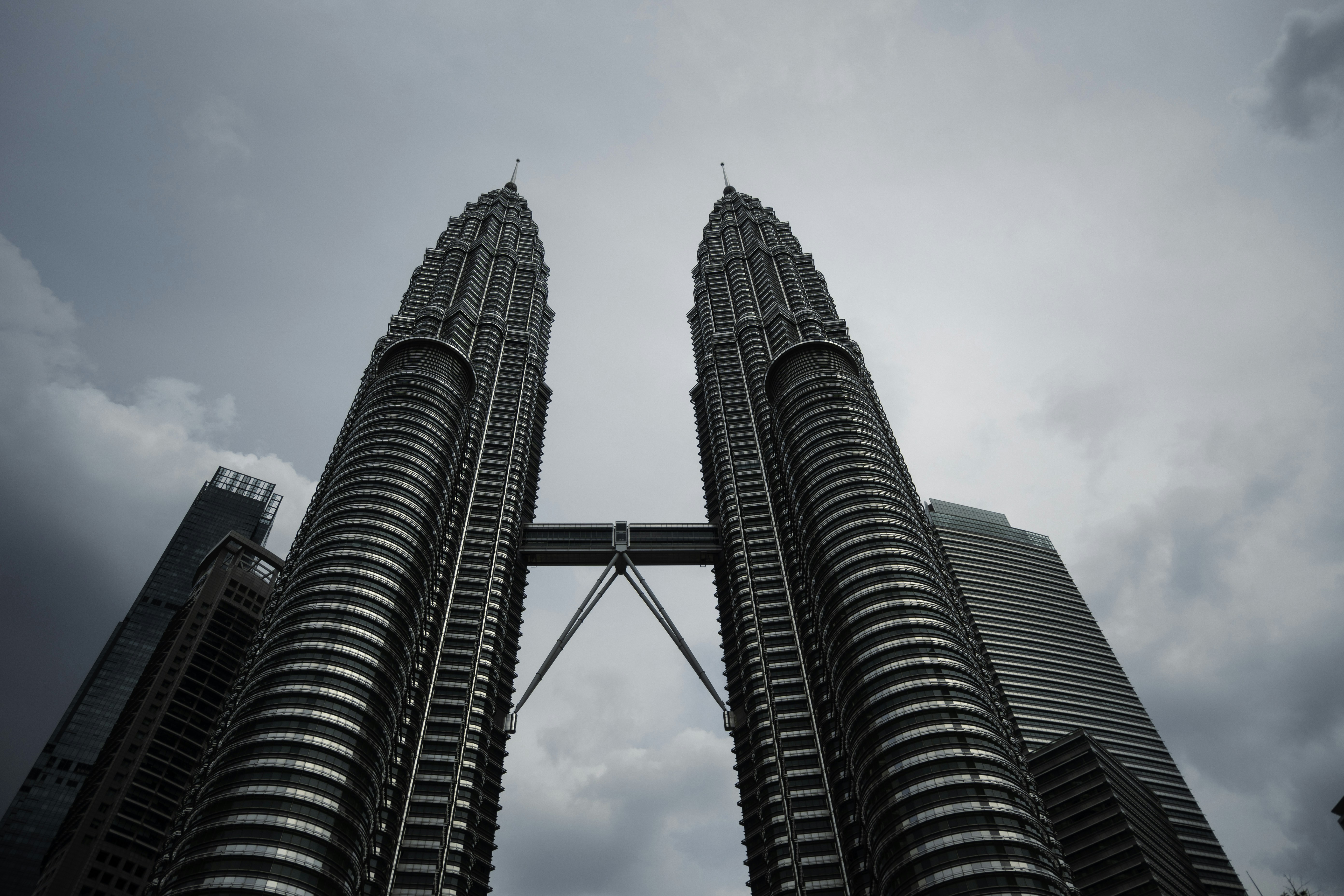Twin skyscrapers rise dramatically against a moody sky, showcasing modern architecture with intricate details. The image emphasizes the connection between the towers via a sky bridge.