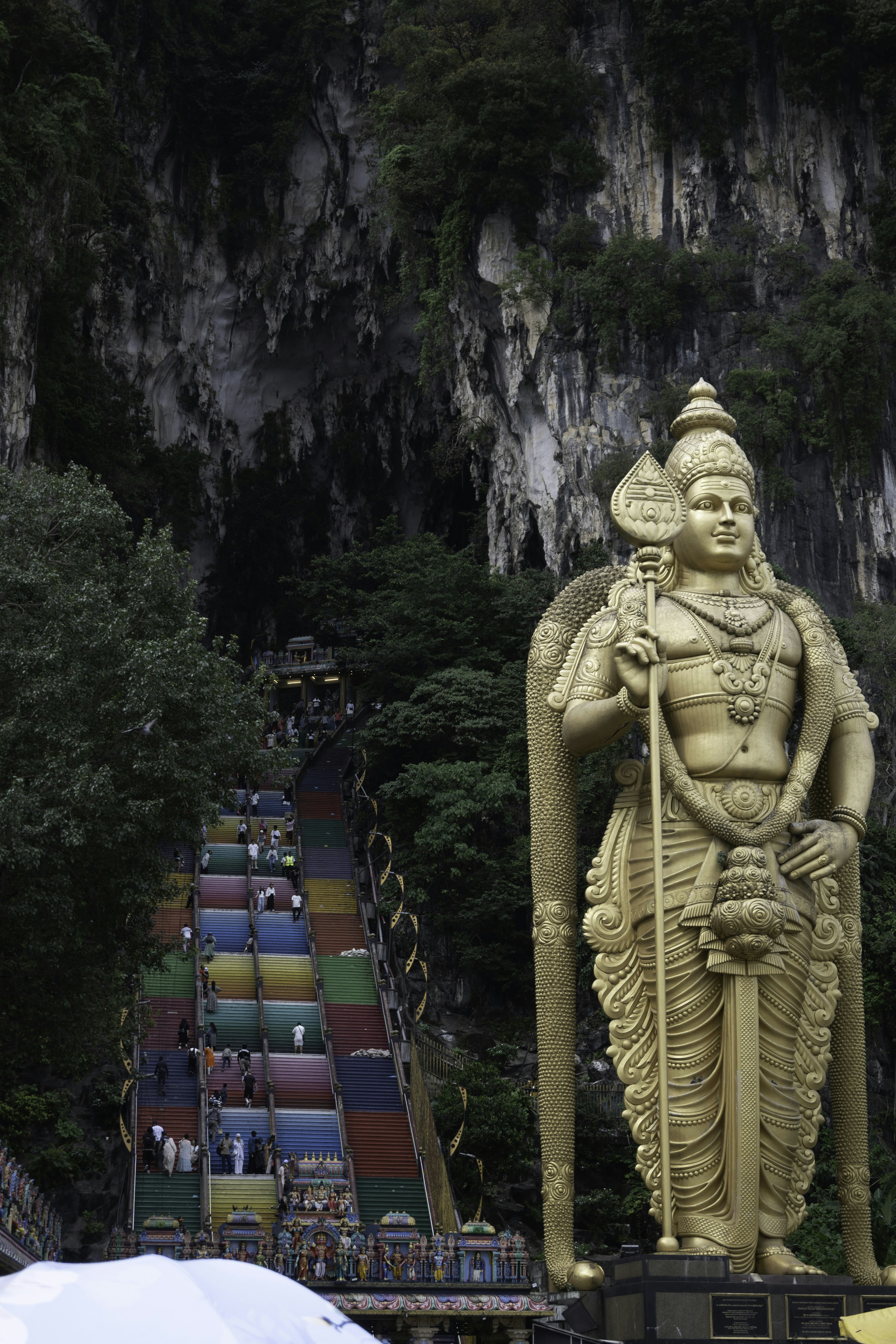 Majestic golden statue of a deity stands at the foot of colorful steps leading into a limestone cave. Lush greenery surrounds the scene, enhancing the spiritual ambiance.