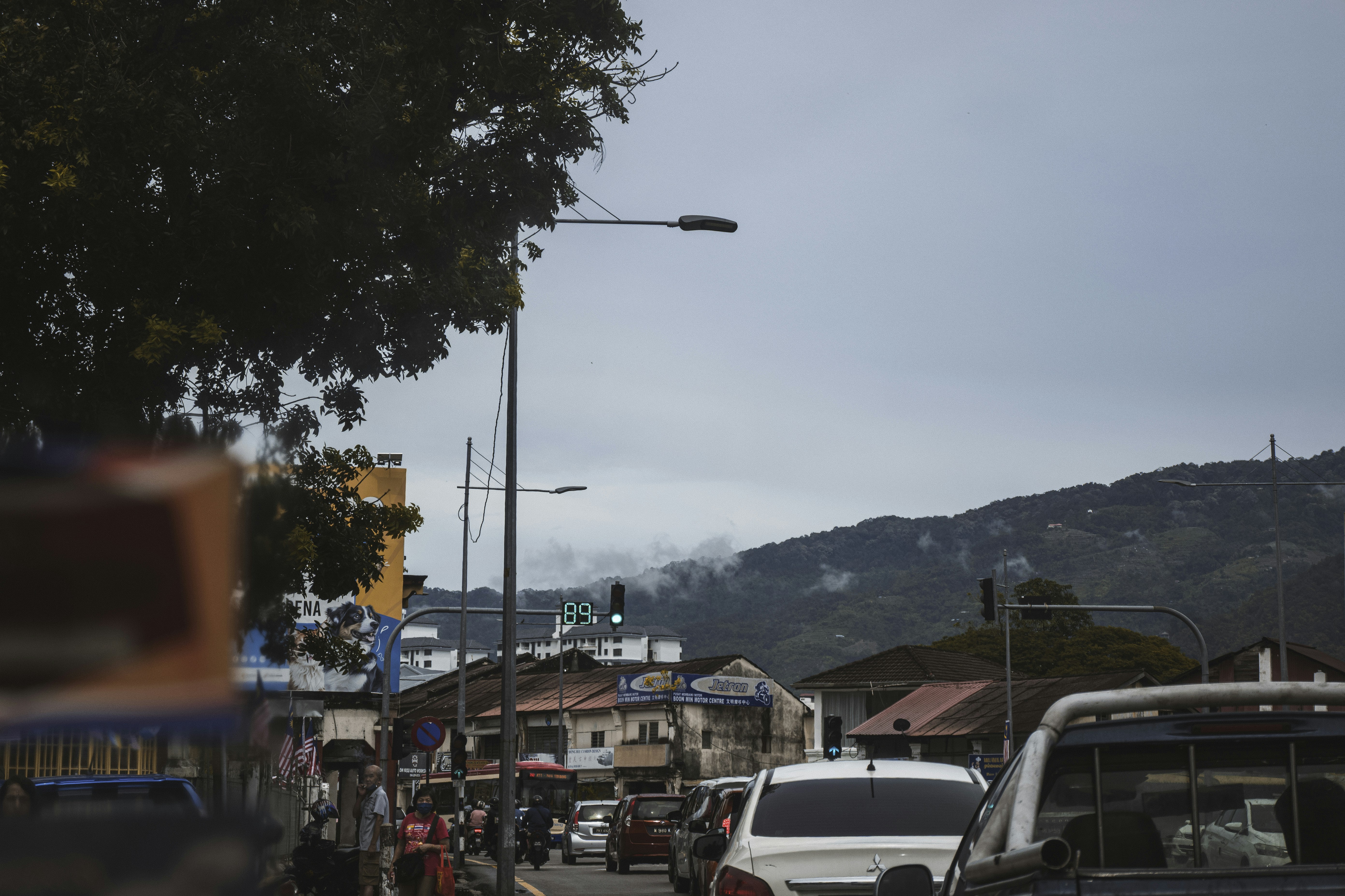 A bustling street scene featuring vehicles and traffic signals, set against a backdrop of rolling hills under a cloudy sky.