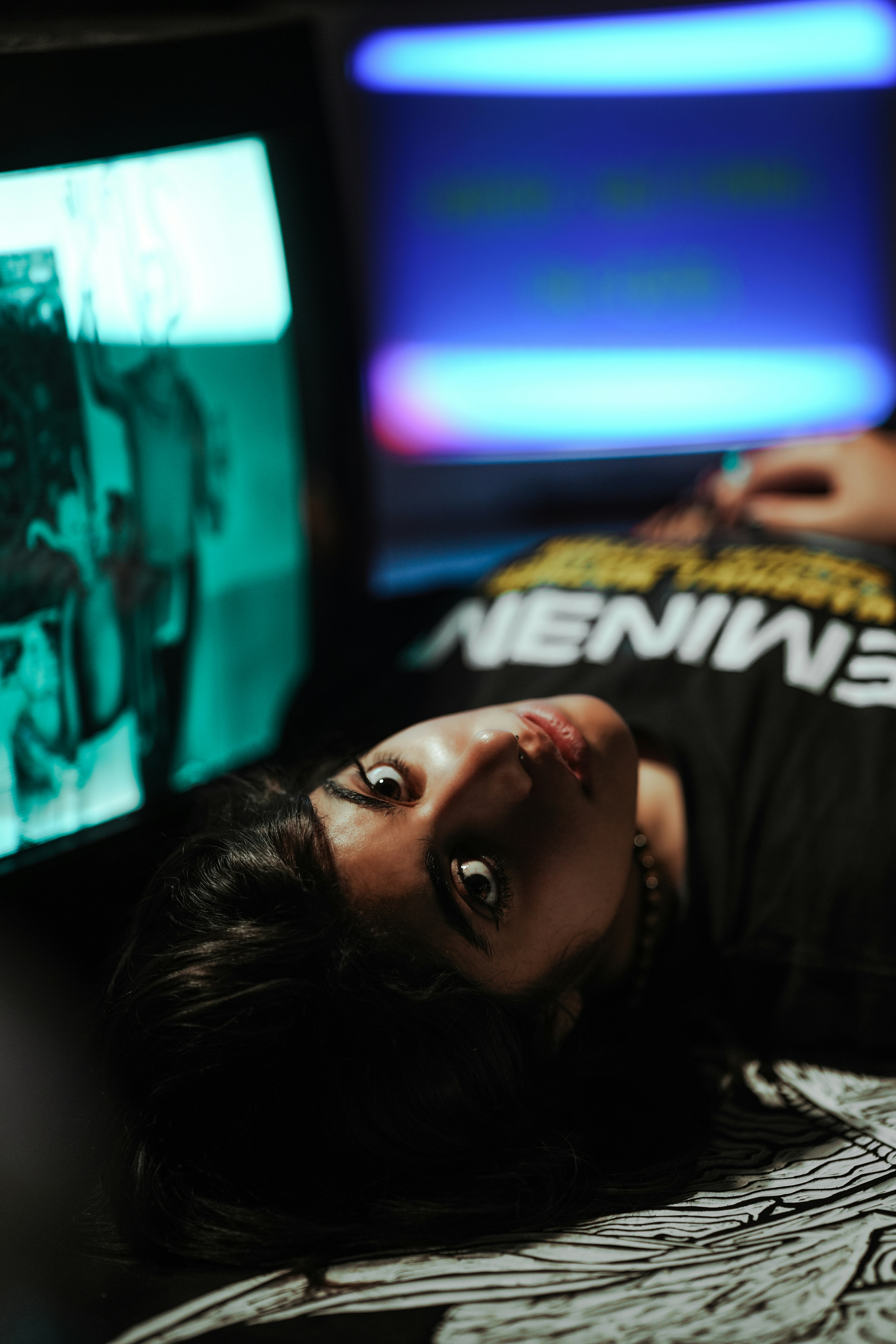 A young person reclines on a patterned surface, gazing intently at the viewer, with a vintage television displaying abstract visuals nearby.