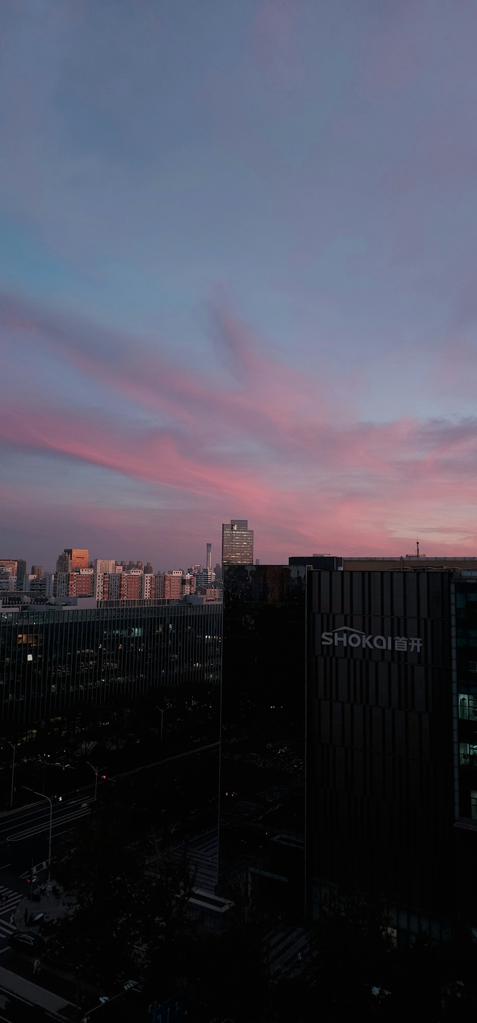City skyline illuminated by soft twilight hues, with clouds painted in shades of pink and blue. Modern buildings reflect the evening light.