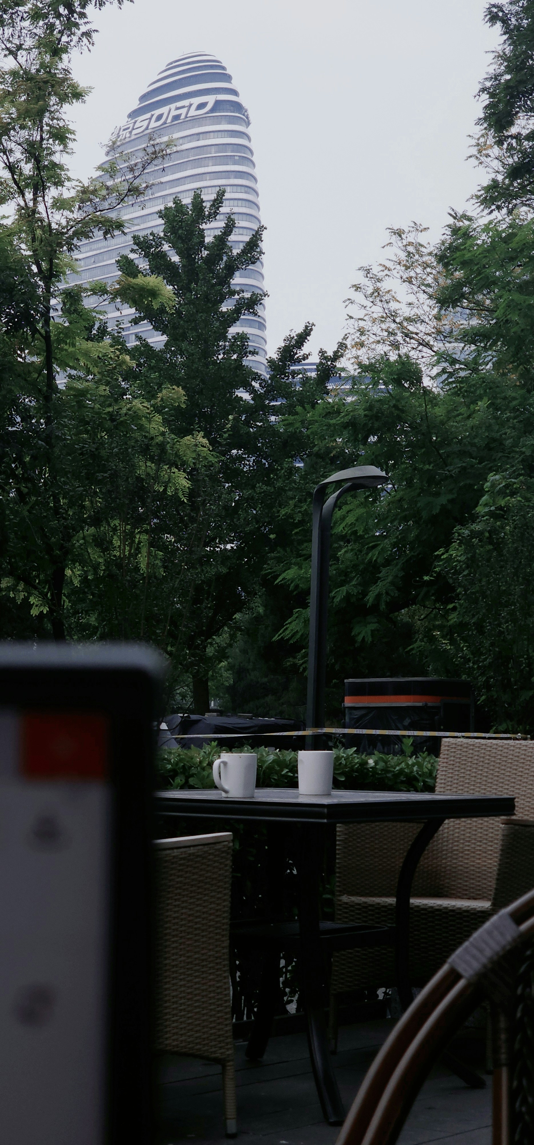 View of a modern skyscraper partially obscured by lush greenery, with a cozy outdoor café setup in the foreground. The scene captures a blend of nature and urban architecture.