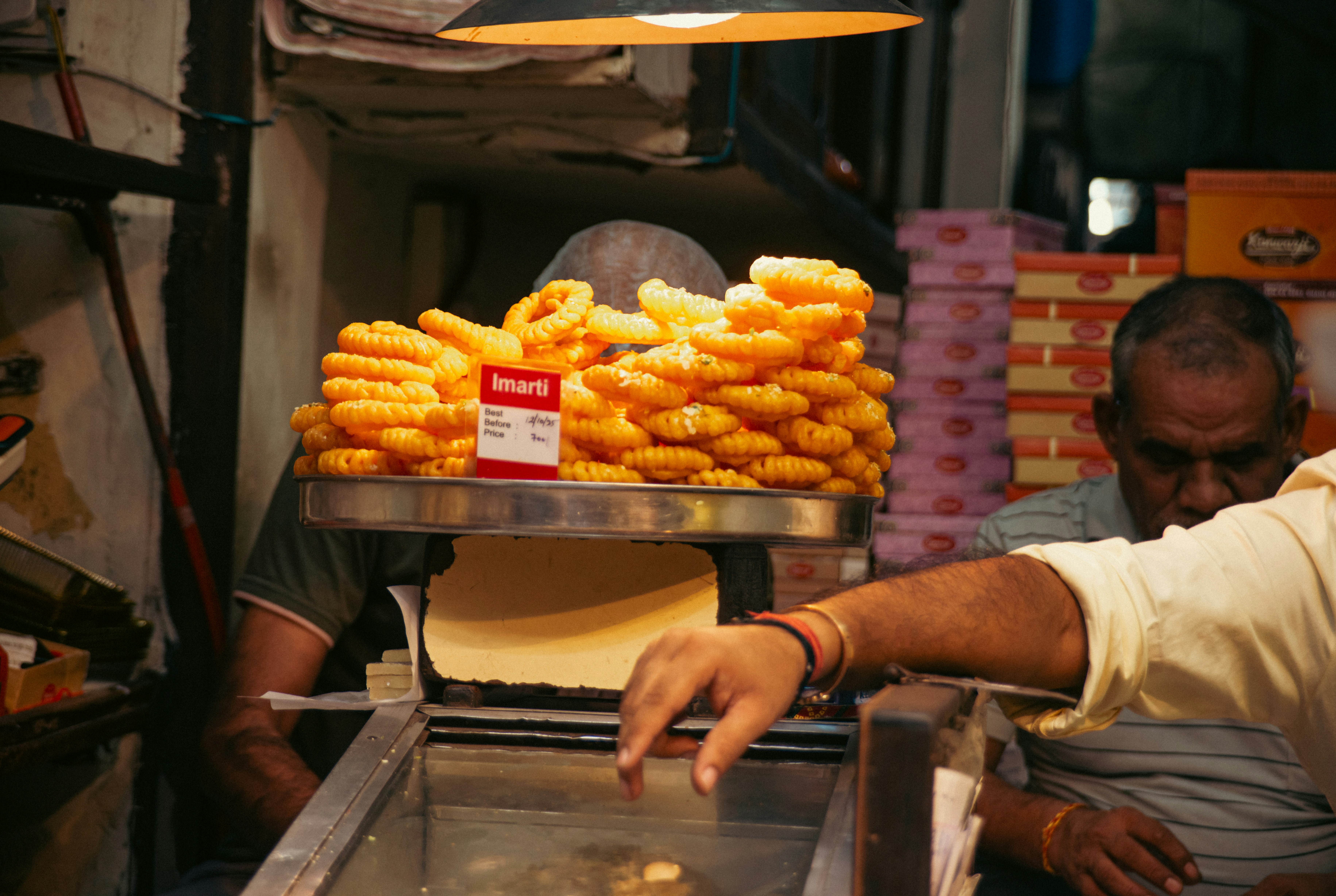 Imarti is a traditional Indian sweet made from deep-fried urad dal batter, which is then soaked in sugar syrup. It is known for its flower-like shape and is different from jalebi because it is made from lentils (urad dal) rather than all-purpose flour (maida). It is a popular, crispy-on-the-outside and juicy-on-the-inside treat often served warm. | Sweet fried dough rings stacked at a market stall.