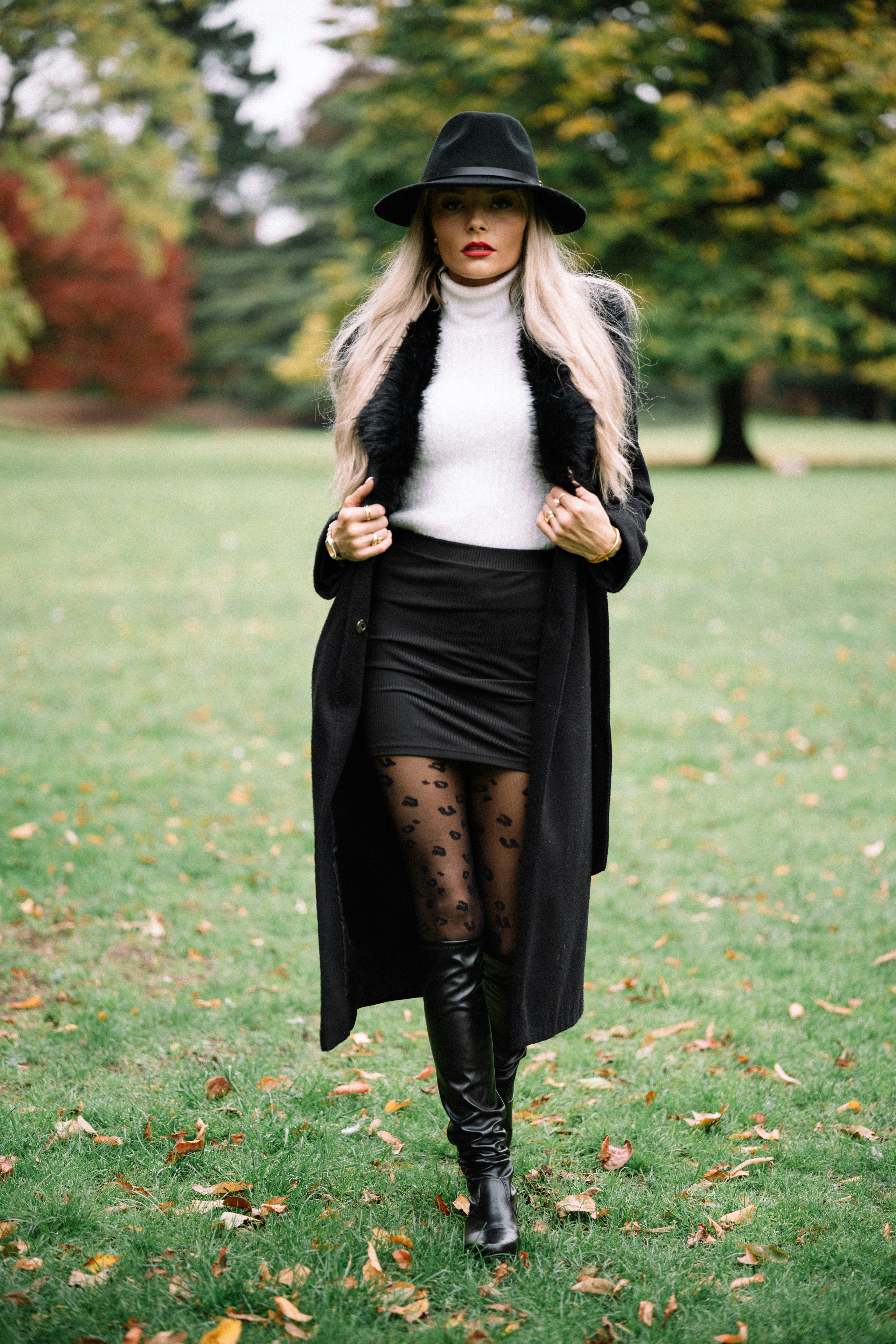 A fashionable woman in a black coat and hat stands confidently on a grassy field adorned with fallen leaves.