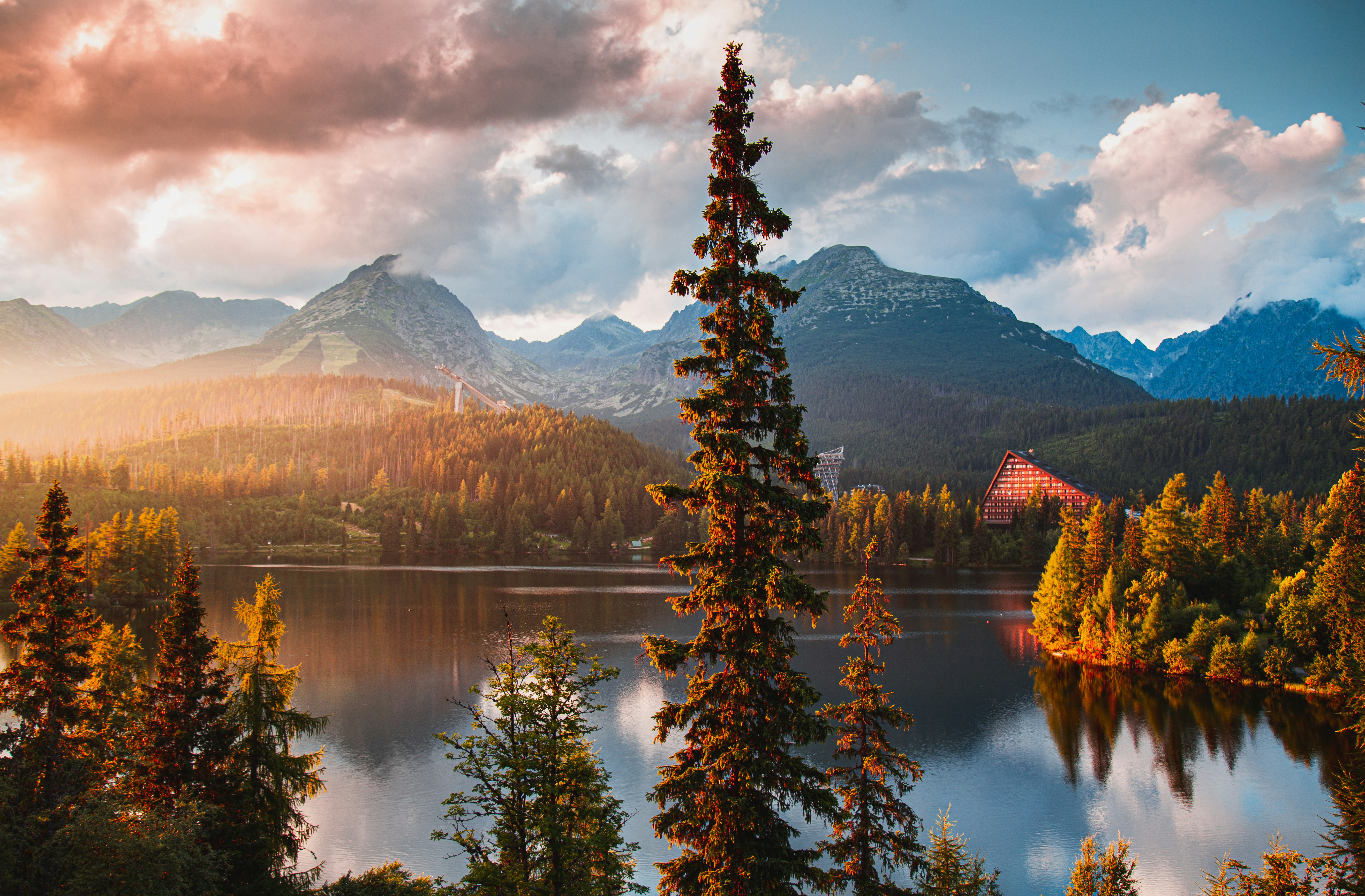 Lush pine trees frame a tranquil lake reflecting majestic mountains under a colorful sky at sunset.