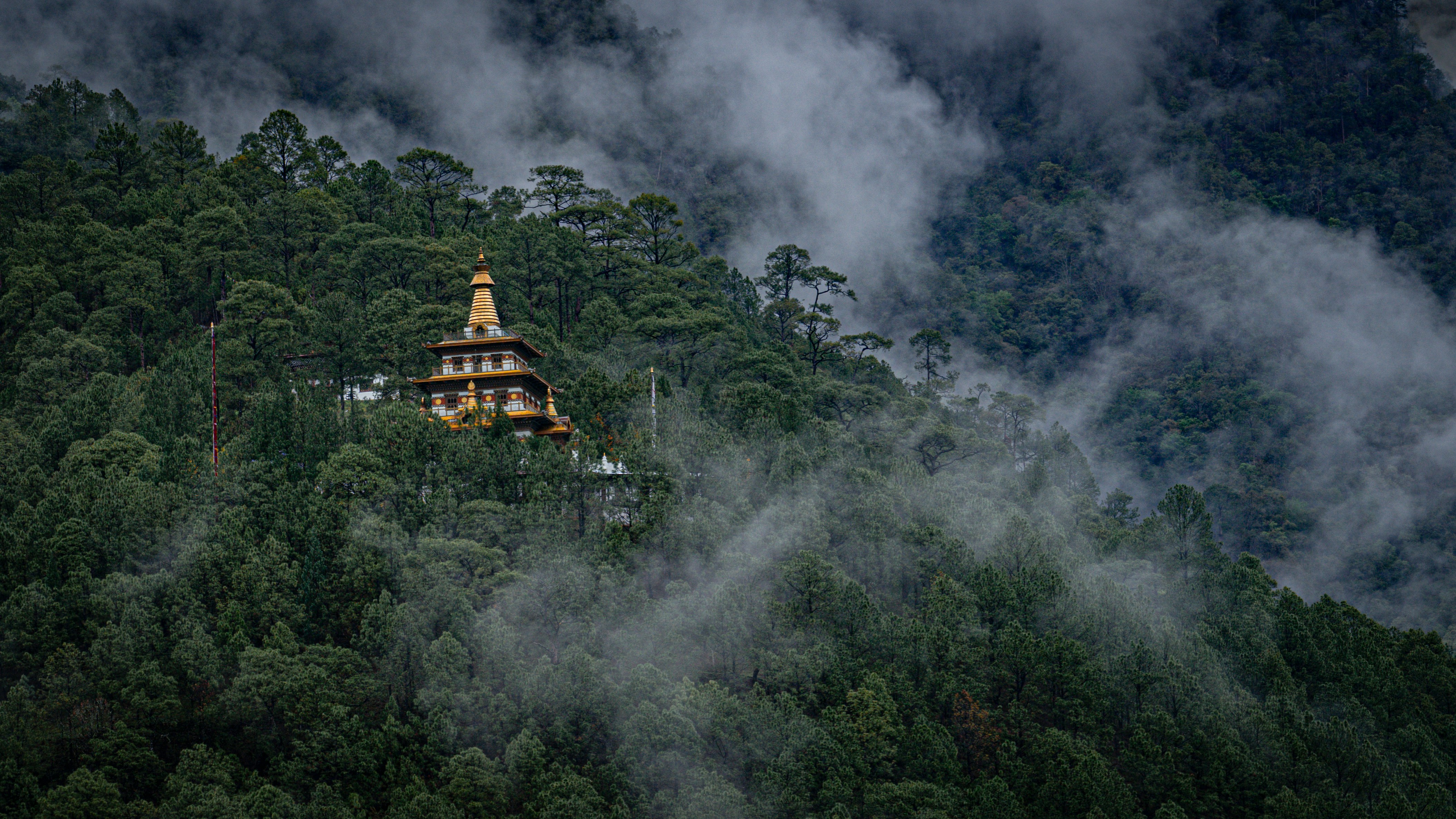 A serene monastery perched on a hillside, enveloped by dense green trees and ethereal fog. The golden spire stands out against the moody backdrop.