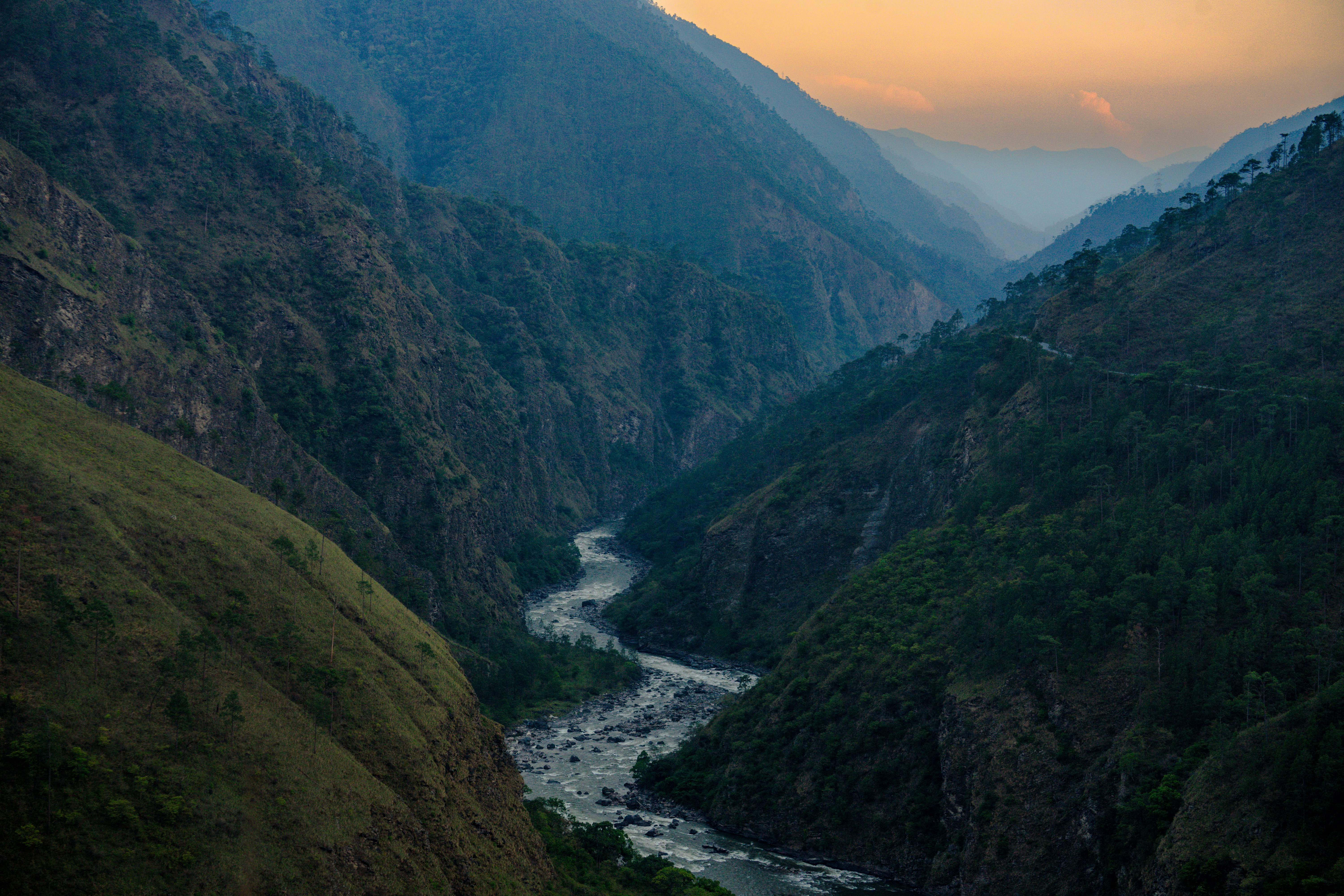 A meandering river flows through steep, rugged valleys, framed by lush greenery and distant mountain silhouettes at sunset.