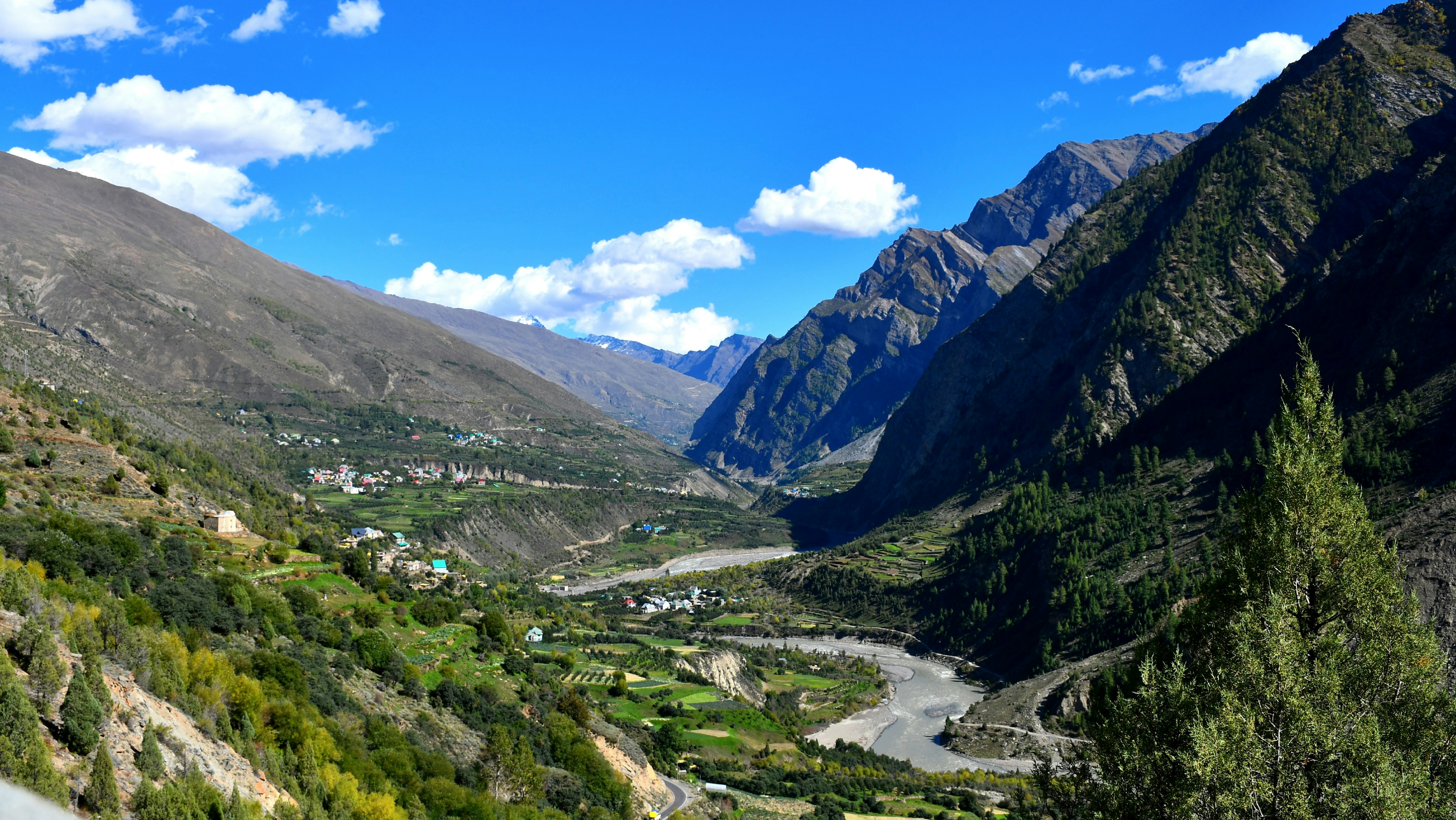 Lush valley nestled between towering mountains, with a winding river and scattered villages under a bright blue sky dotted with clouds.
