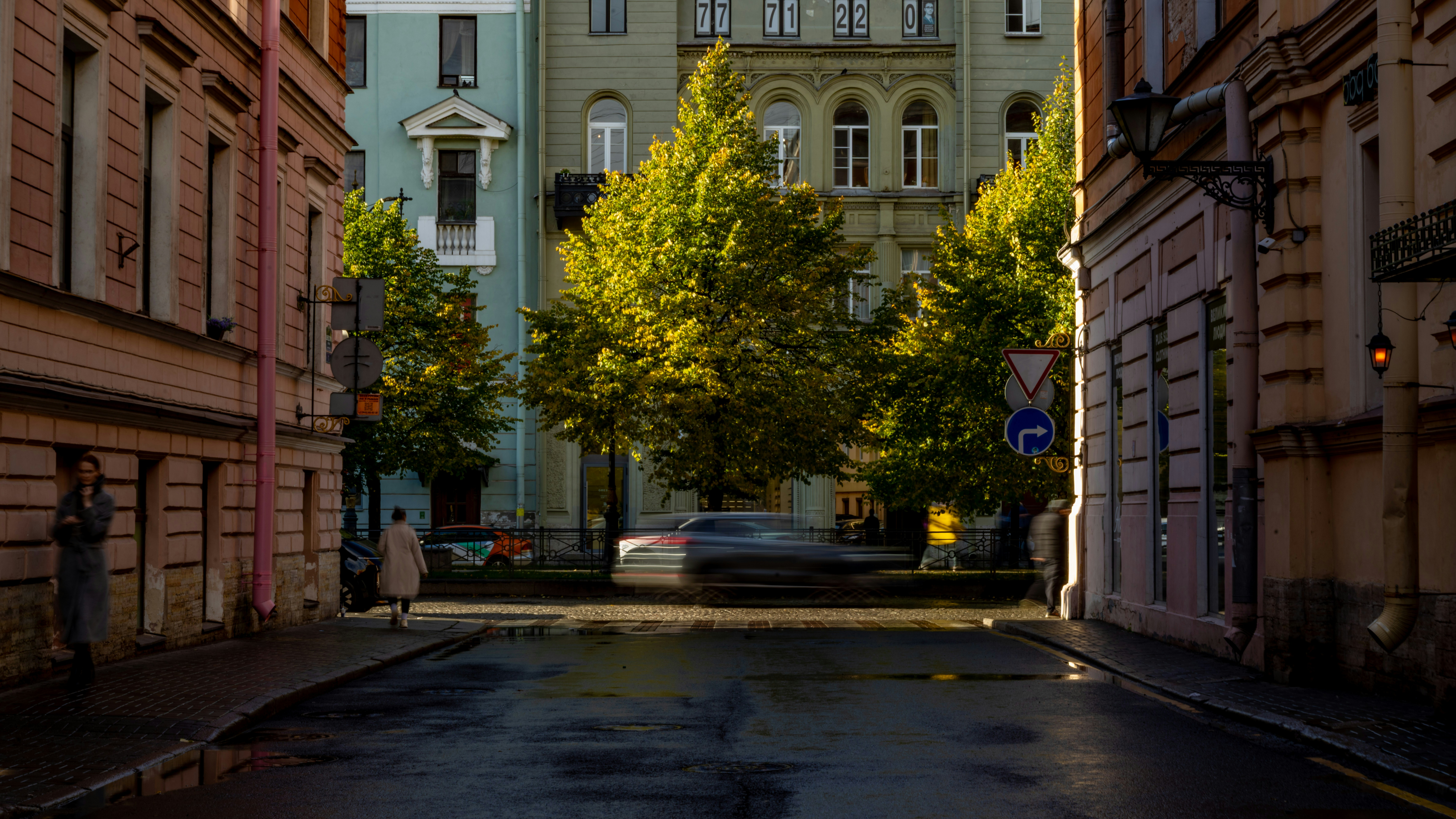 Sunlight filters through trees at the intersection of historic buildings, creating a tranquil urban scene. The reflection on the wet pavement enhances the peaceful atmosphere.