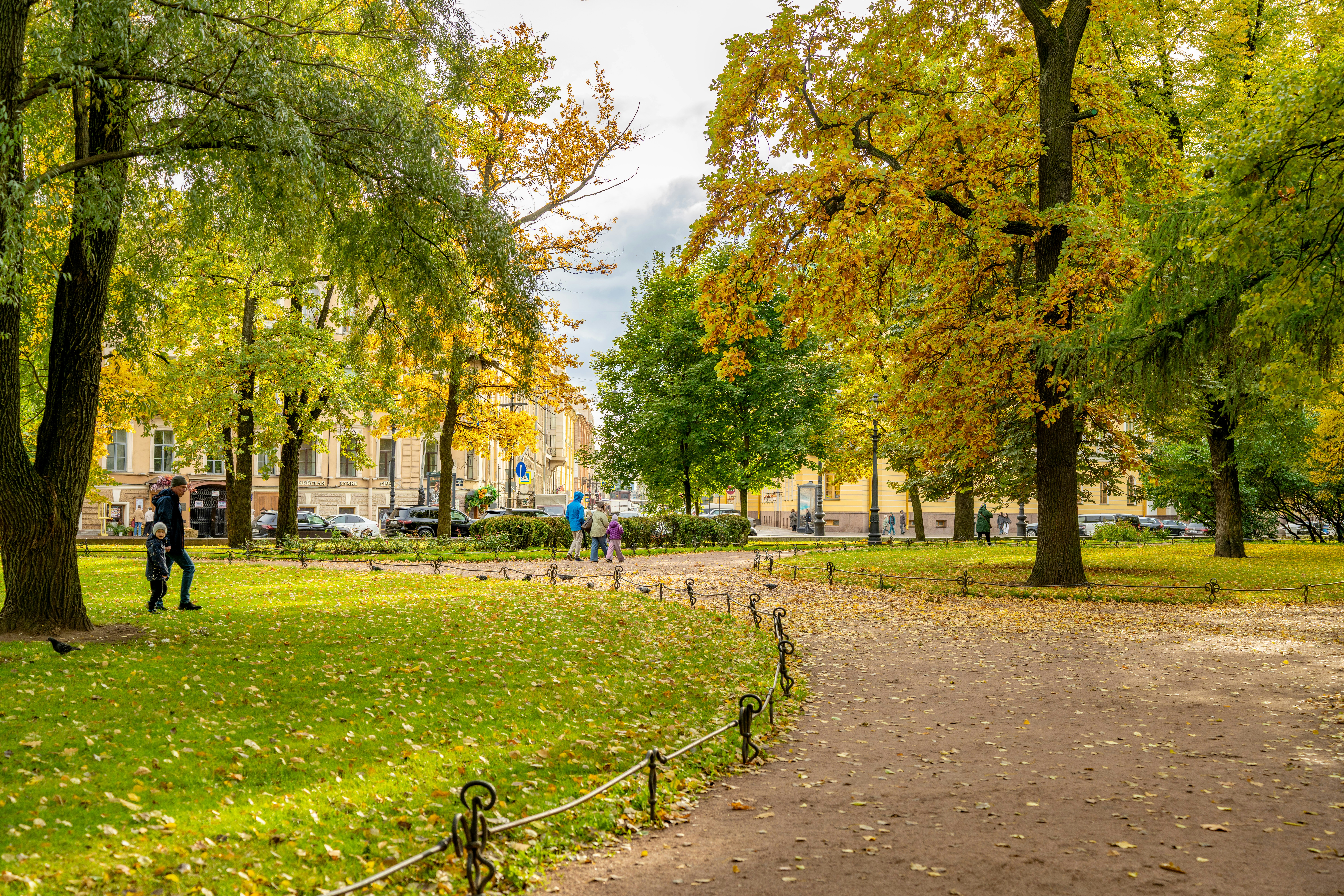 Lush green grass and vibrant autumn foliage frame a serene park scene with people strolling along winding paths. A blend of nature and urban life creates a tranquil atmosphere.