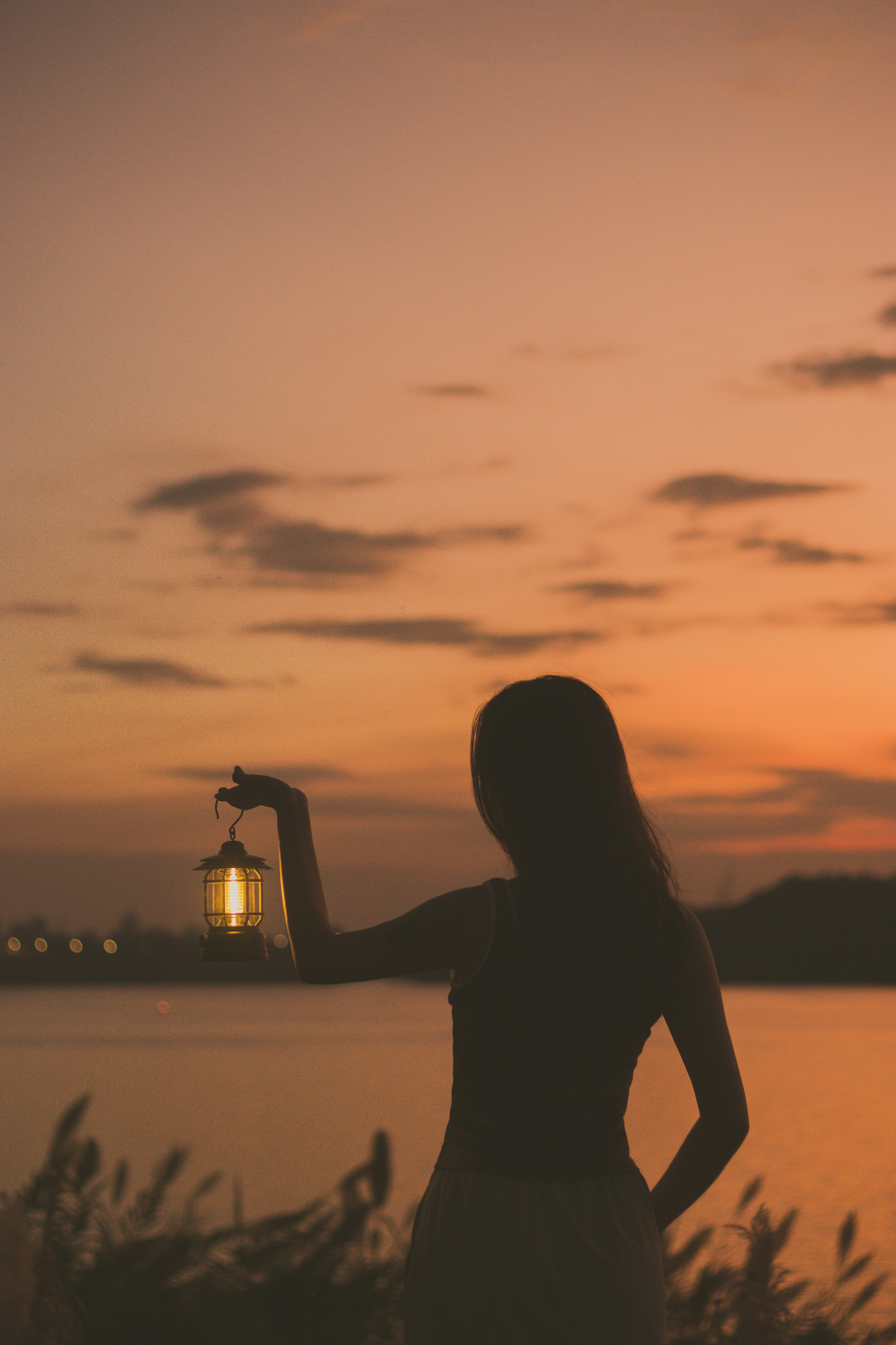 Silhouette of a woman holding a lantern against a vibrant sunset over a calm water body.