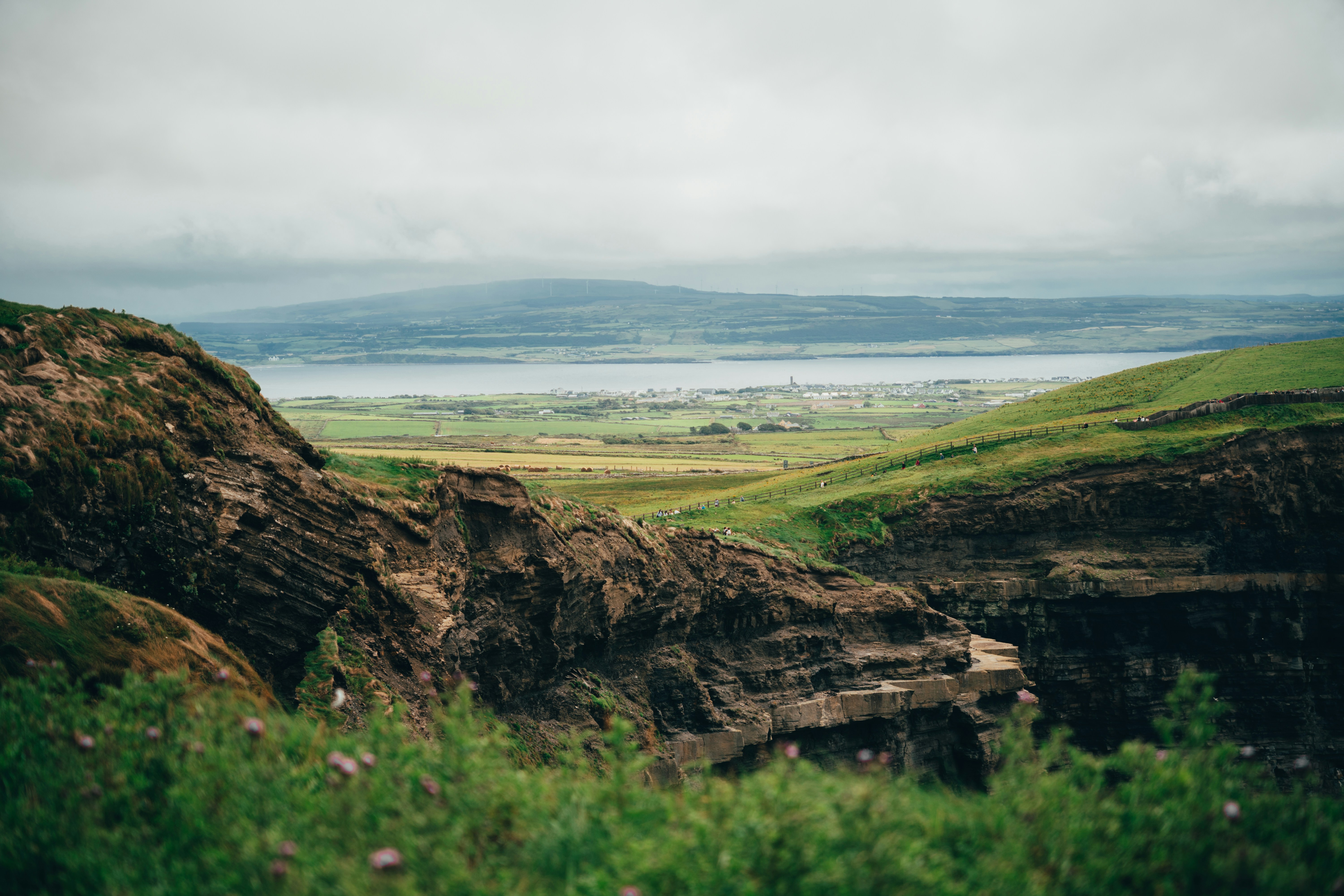 Lush green hills meet rugged cliffs overlooking a tranquil bay, with distant farmland stretching into the horizon.