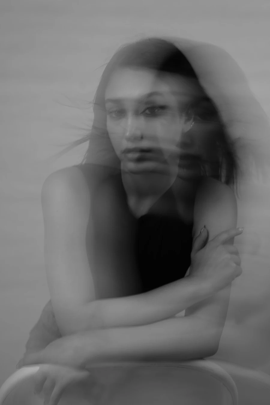 Black-and-white portrait of a woman with motion blur and layered ghosting effects, seated with her arms crossed. Her face remains the central focus against a soft, minimal background.