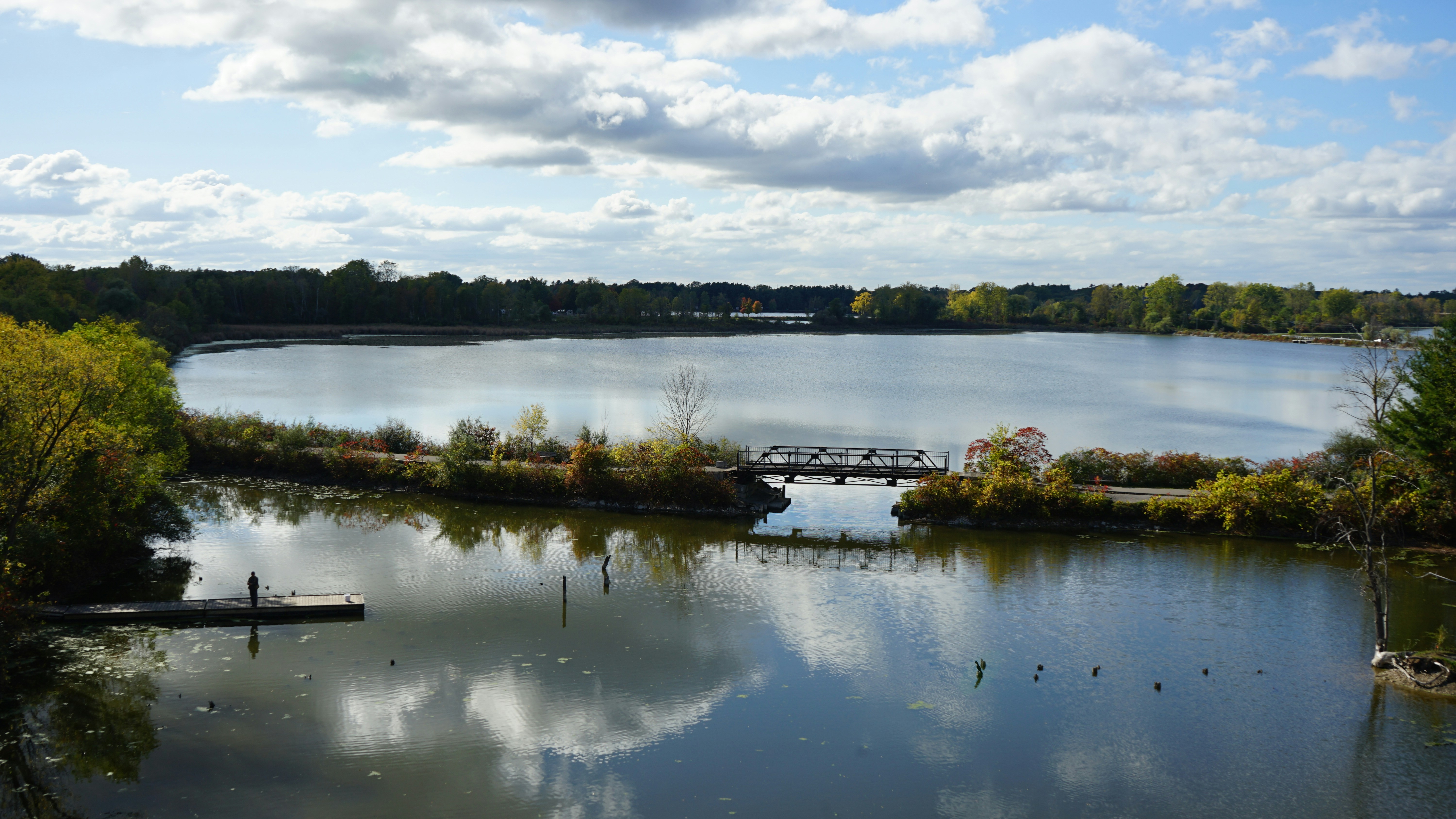 Serene lakeside scene showcasing vibrant autumn foliage and a calm water surface reflecting the sky. A wooden dock extends into the water, enhancing the peaceful ambiance.