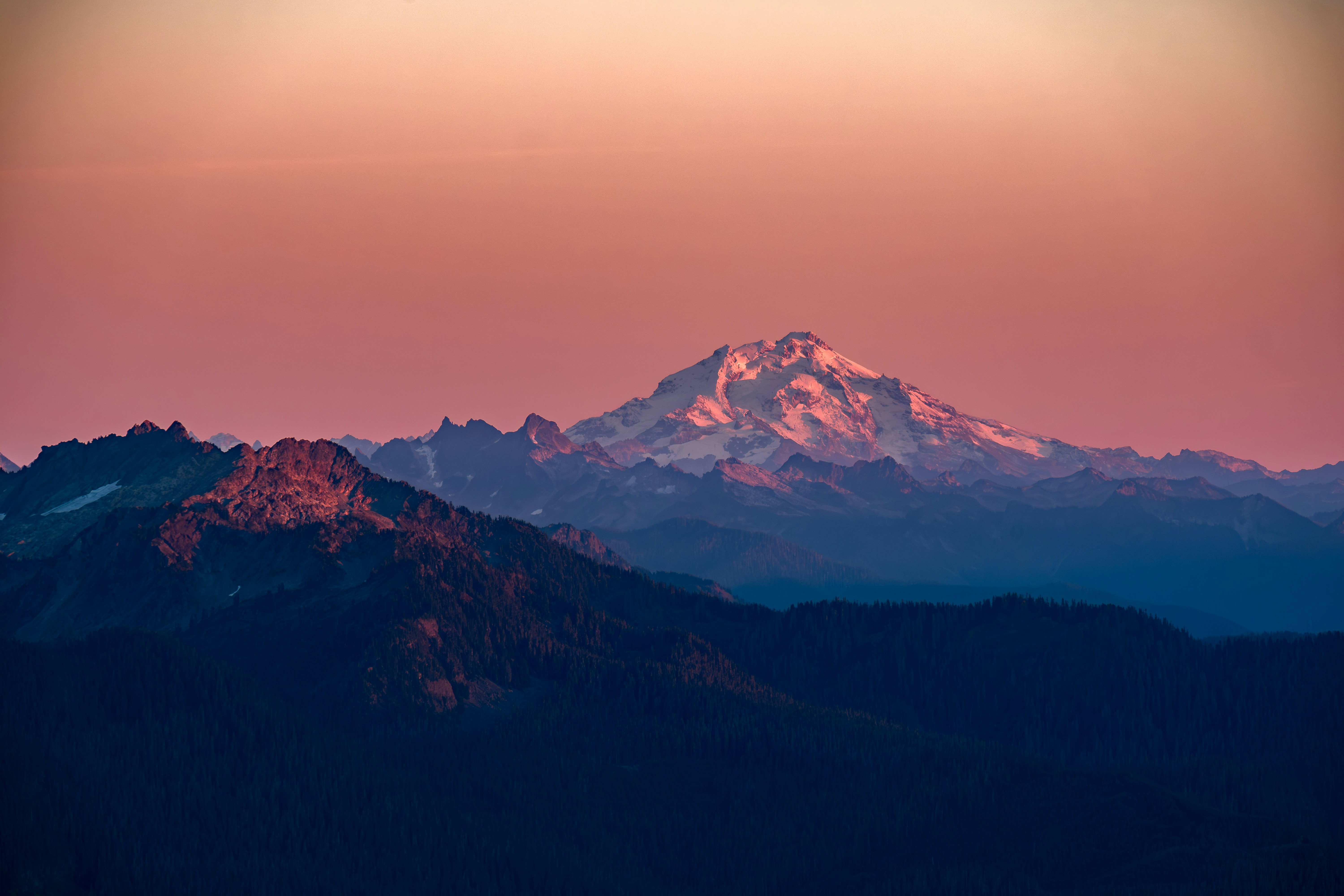 Snow-capped mountain rises above a layered landscape at twilight, bathed in soft hues of pink and purple.