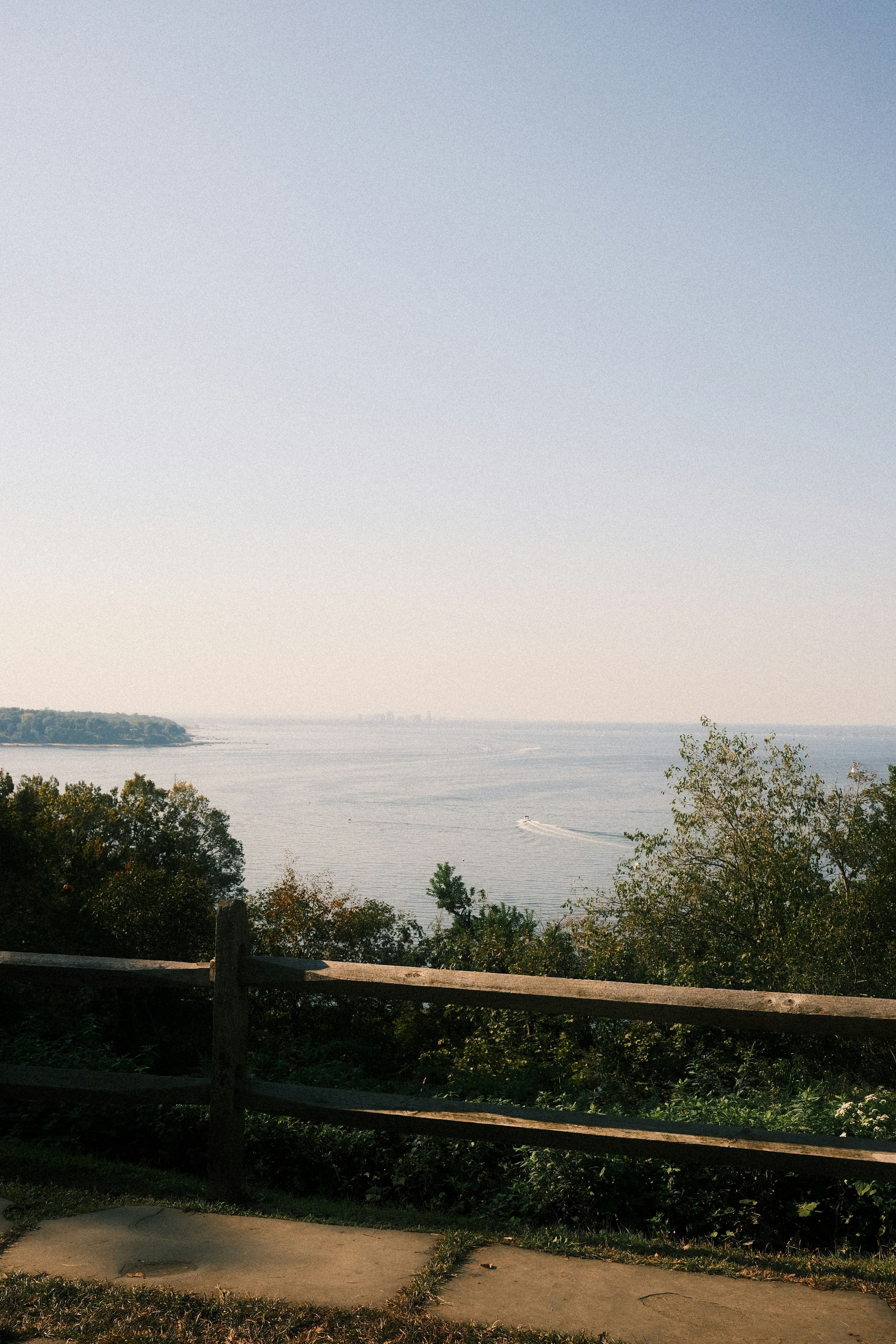 Serene lakeside view with a distant city skyline under a clear sky, framed by lush greenery and wooden fencing.