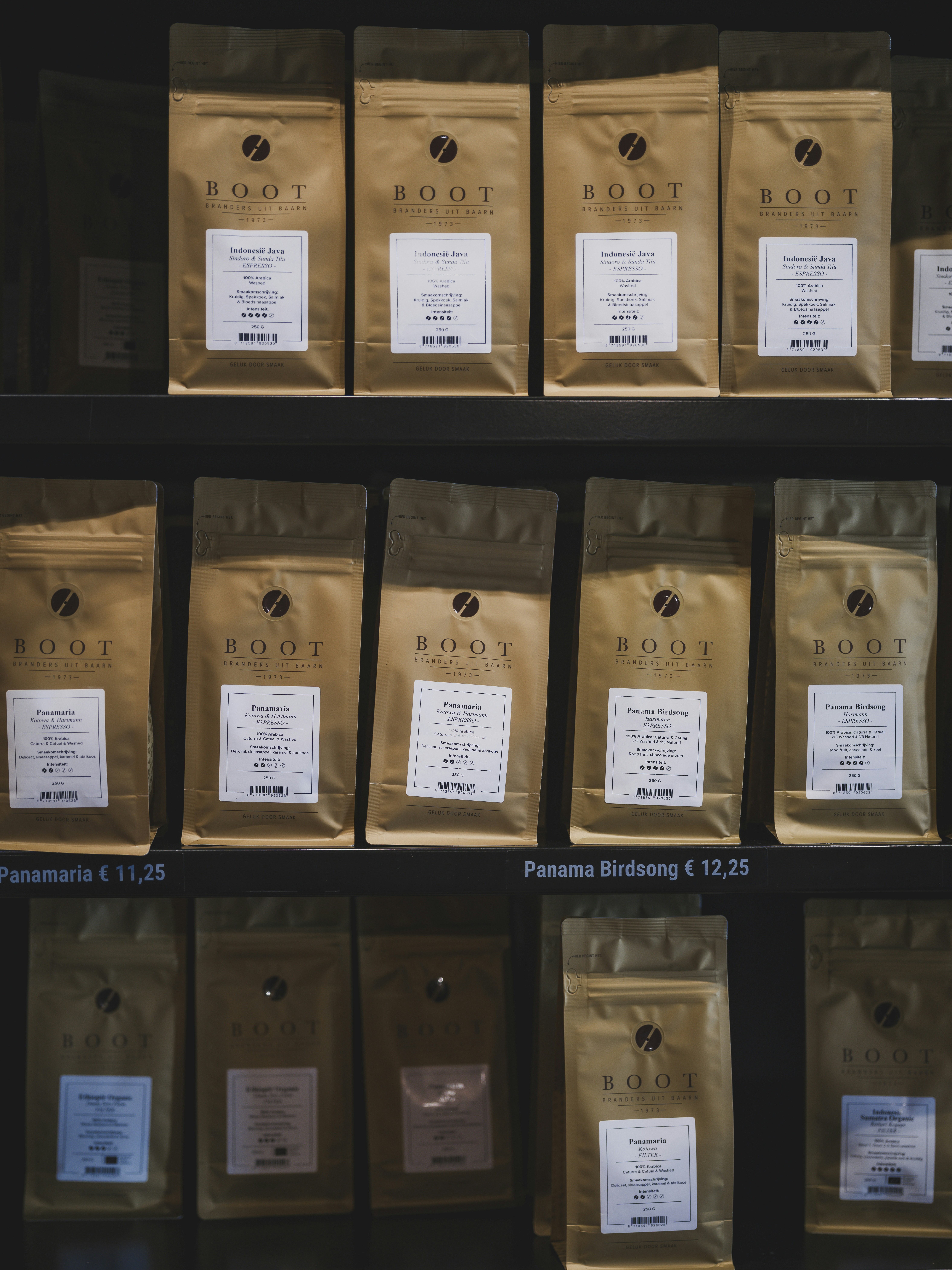 Neatly arranged coffee bags on a shelf, showcasing various blends and origins with elegant gold packaging. Each bag features a distinct label and price tag.