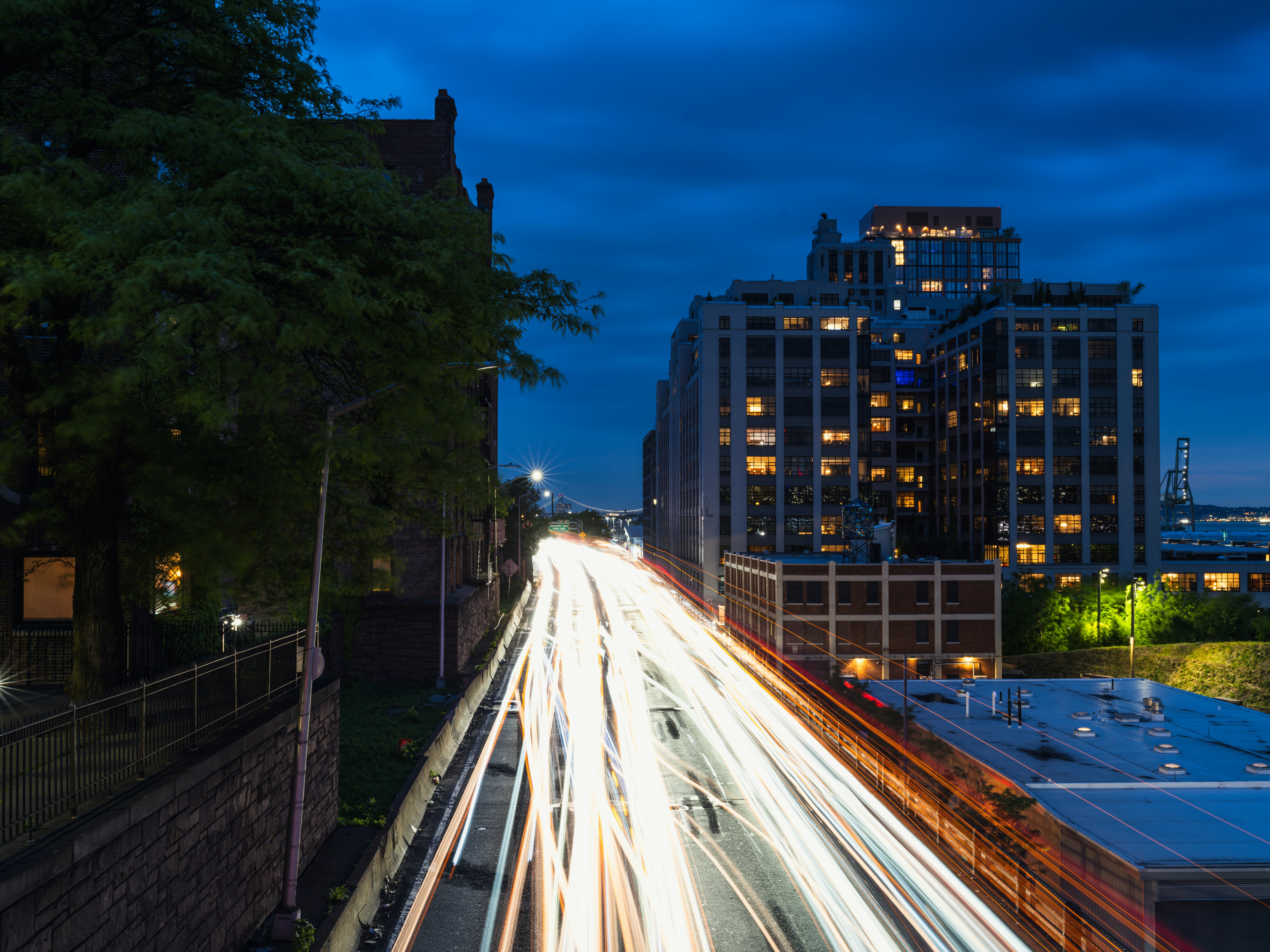 Long exposure captures the vibrant movement of traffic against a twilight cityscape, highlighting architectural details and lush greenery.