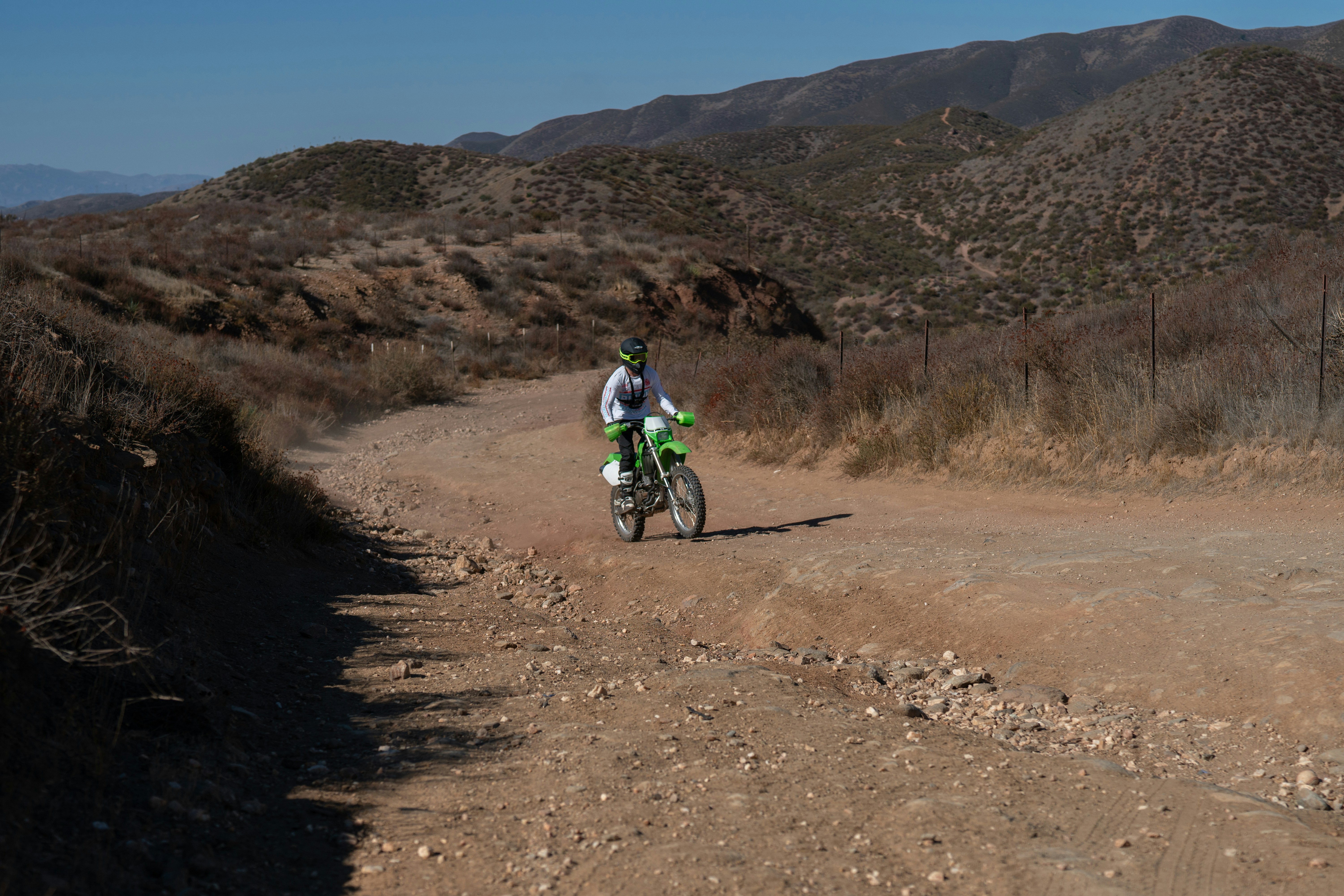 Motocross rider navigating a rugged dirt path through arid hills under a clear blue sky.