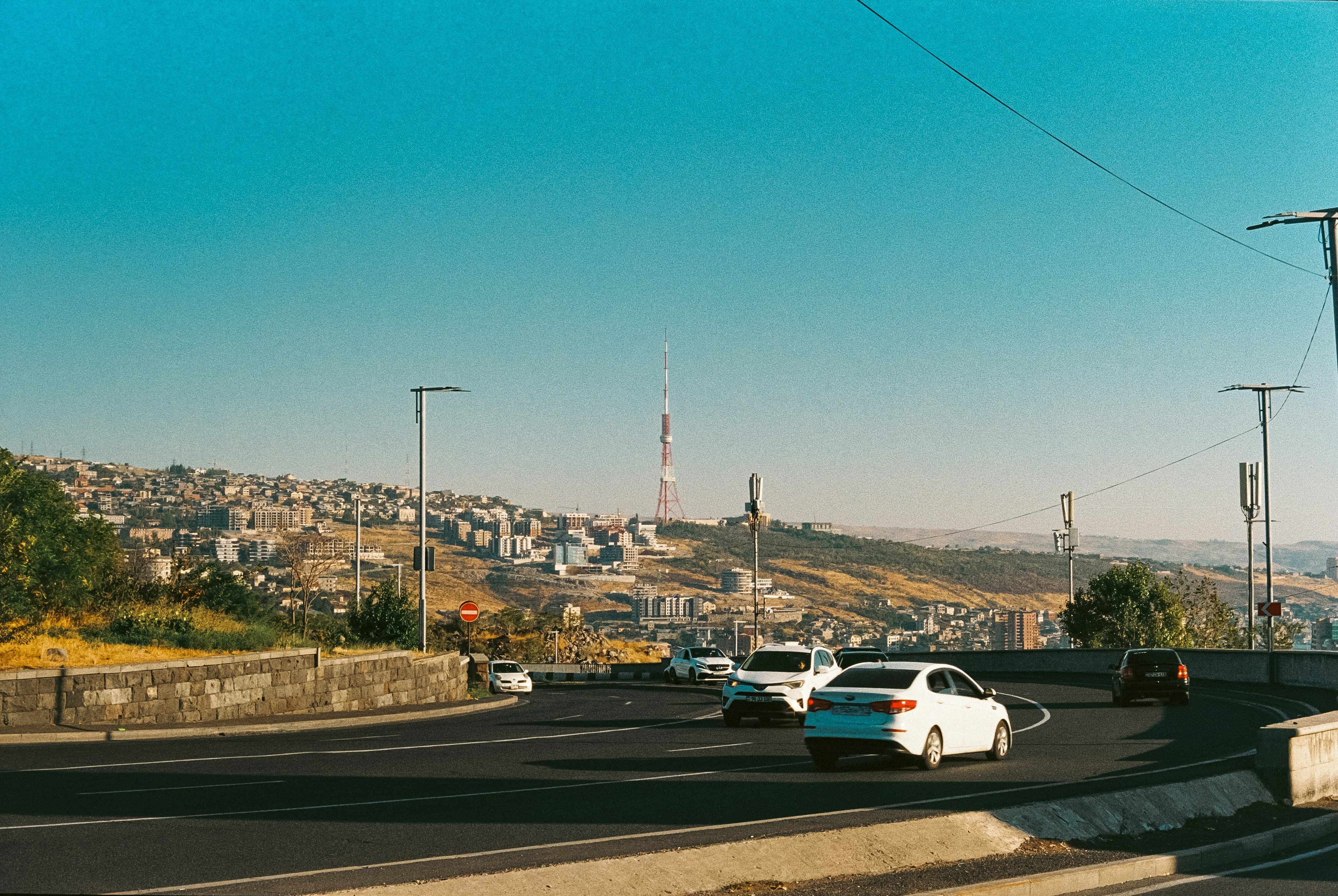 A winding road curves through a vibrant cityscape, showcasing a blend of modern architecture and natural hills, with a prominent telecommunications tower in the background.