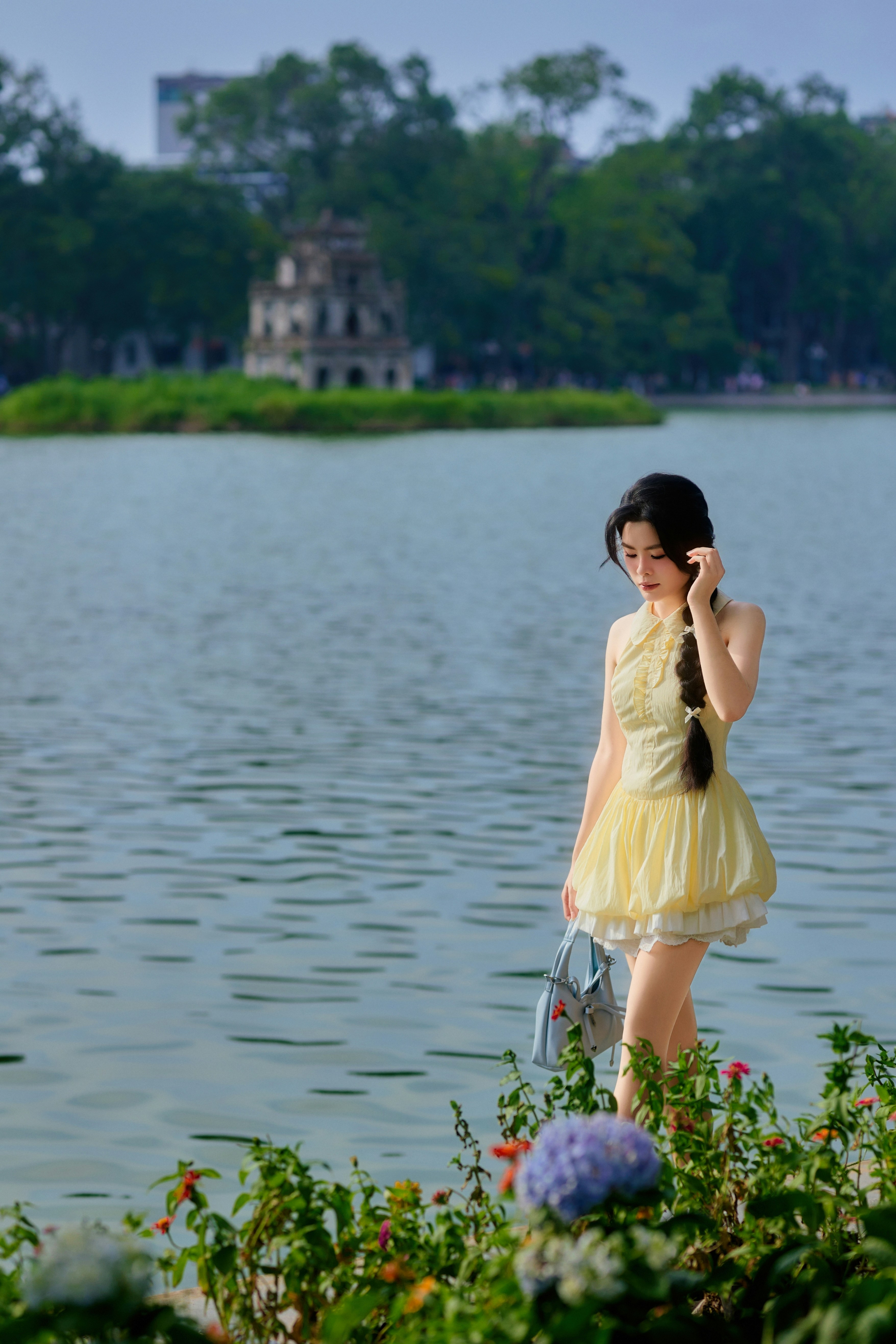 Young woman in a yellow dress walks along the shore of a tranquil lake, surrounded by vibrant flowers and greenery. The distant pagoda adds a cultural touch to the serene landscape.