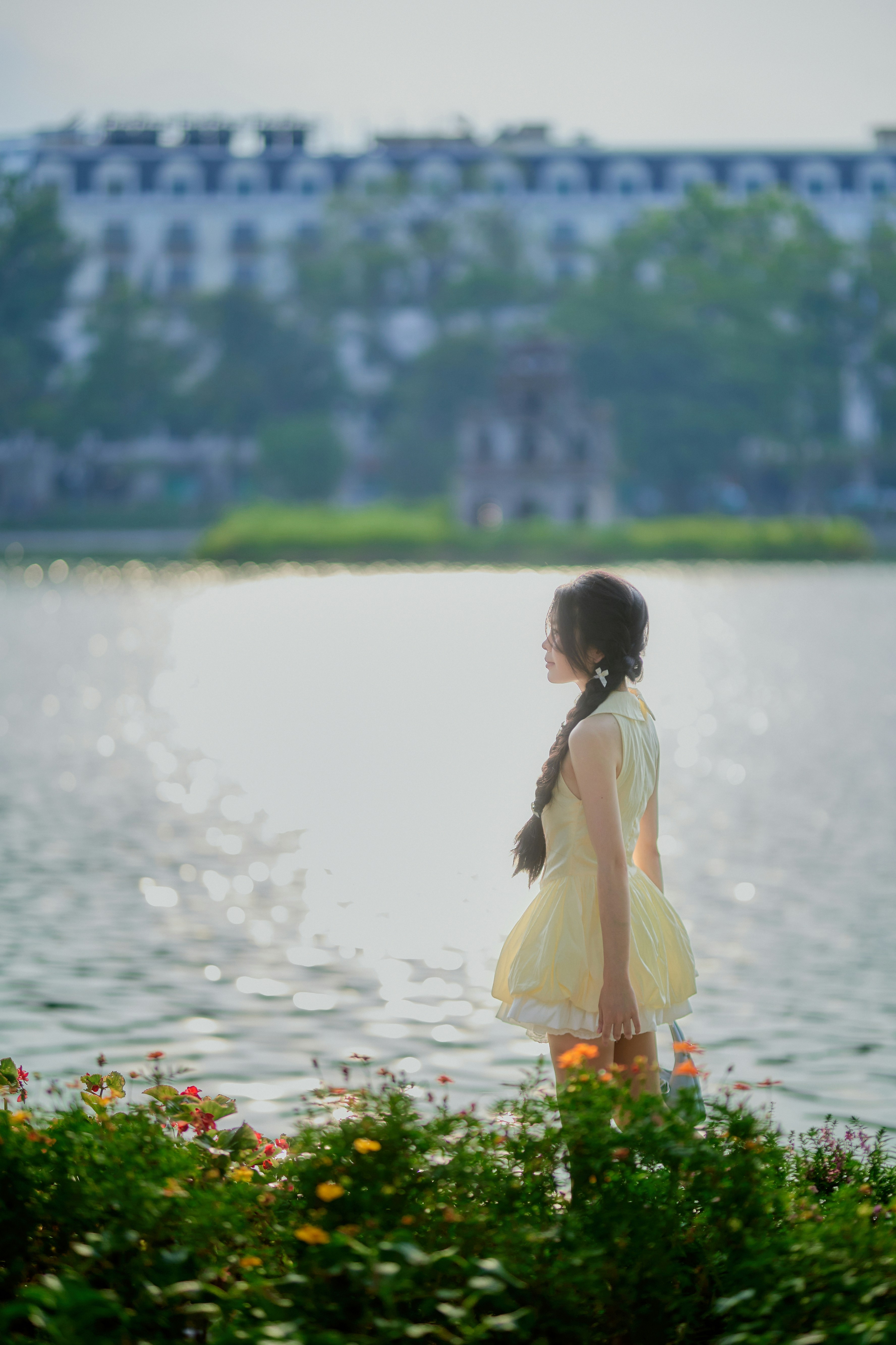 Young woman in a yellow dress stands by a shimmering lake, surrounded by colorful flowers, as sunlight dances on the water&#x27;s surface.