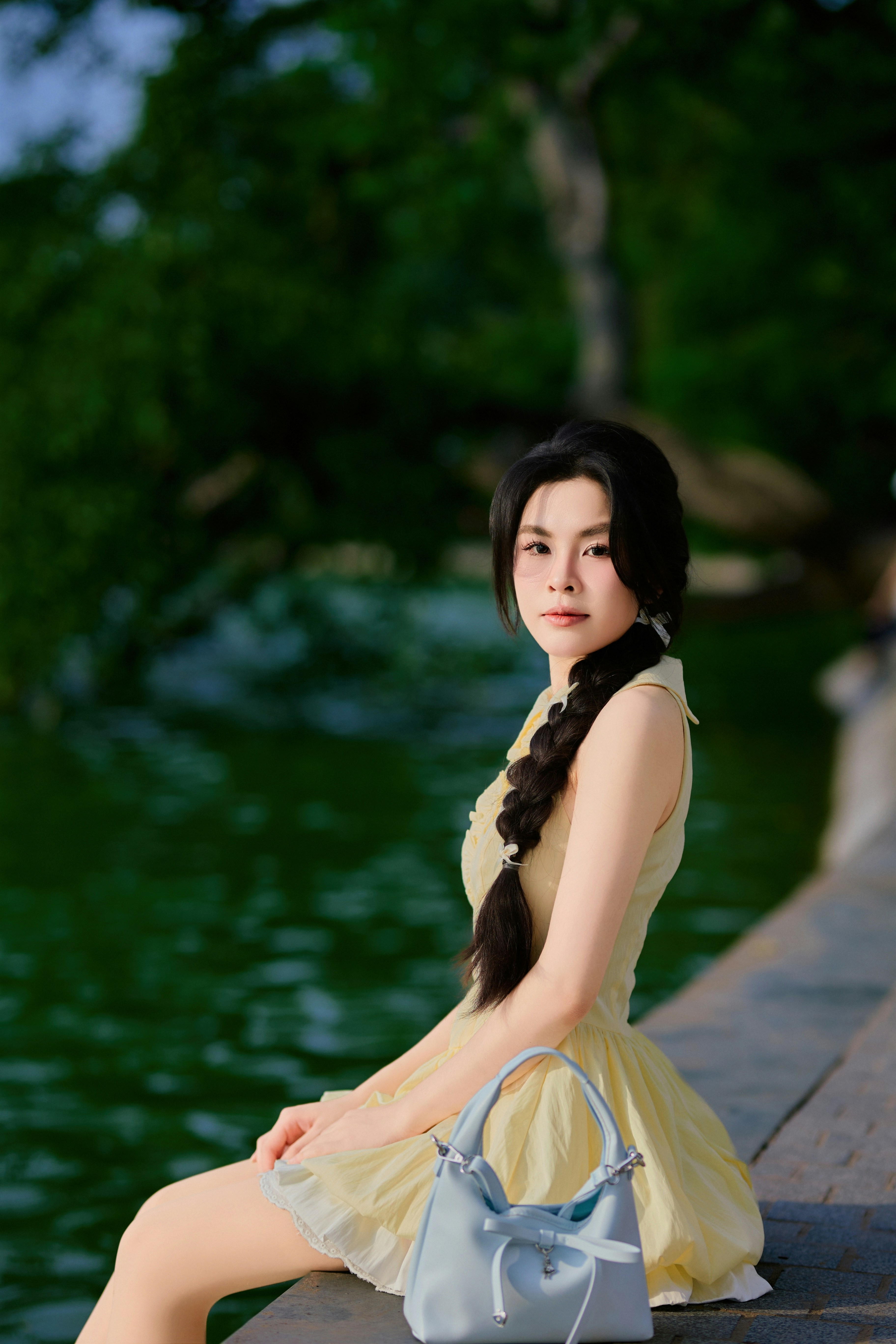 A young woman in a yellow dress sits gracefully by a tranquil lake, her braided hair gently cascading over her shoulder. The lush greenery and calm water reflect a peaceful summer day.