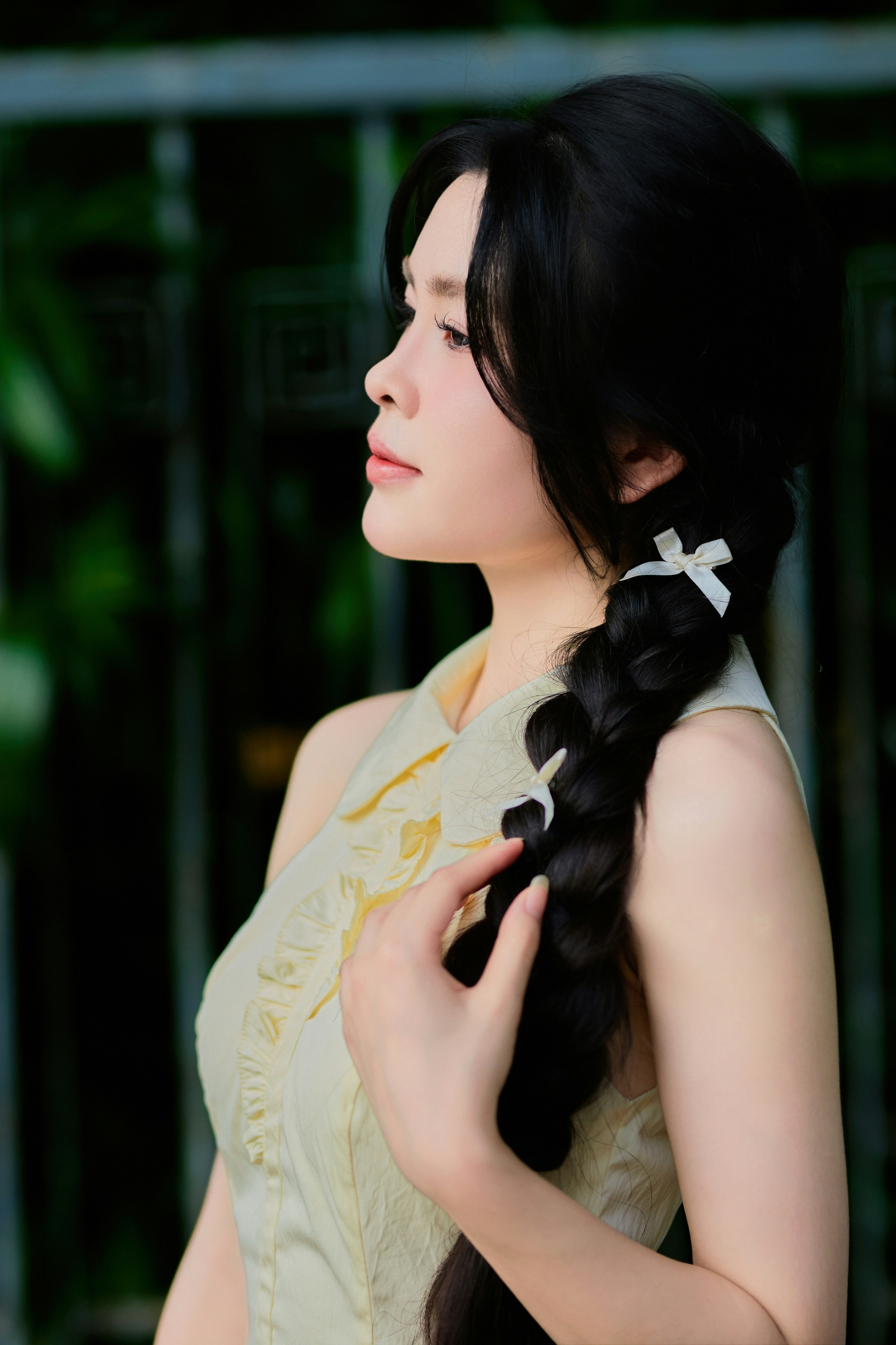 A young woman with a braided hairstyle adorned with ribbons gazes thoughtfully, surrounded by lush greenery.