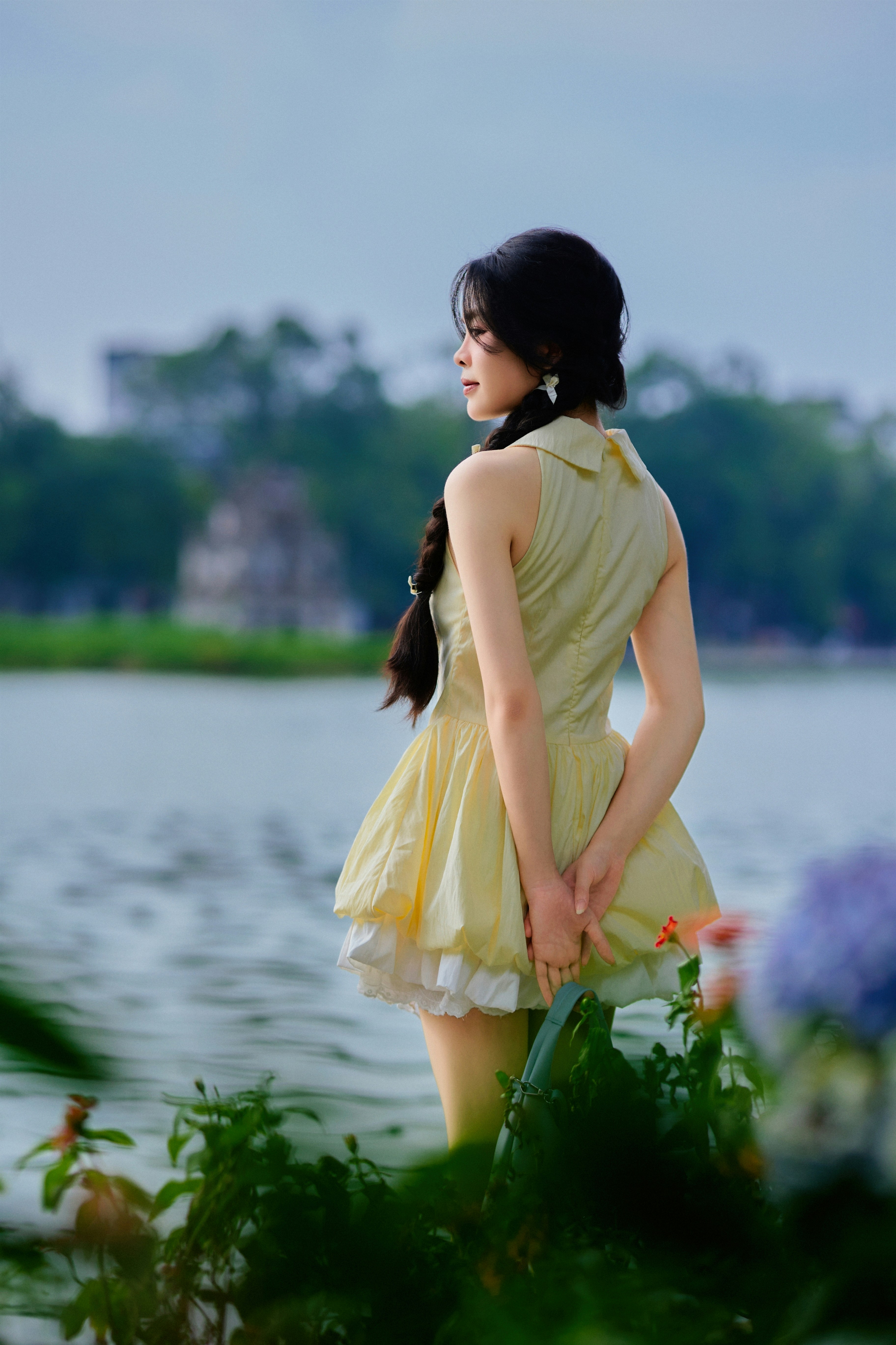 Young woman in a soft yellow dress stands by a serene lake, with a gentle breeze ruffling her hair. The scene is framed by lush greenery and blooming flowers.