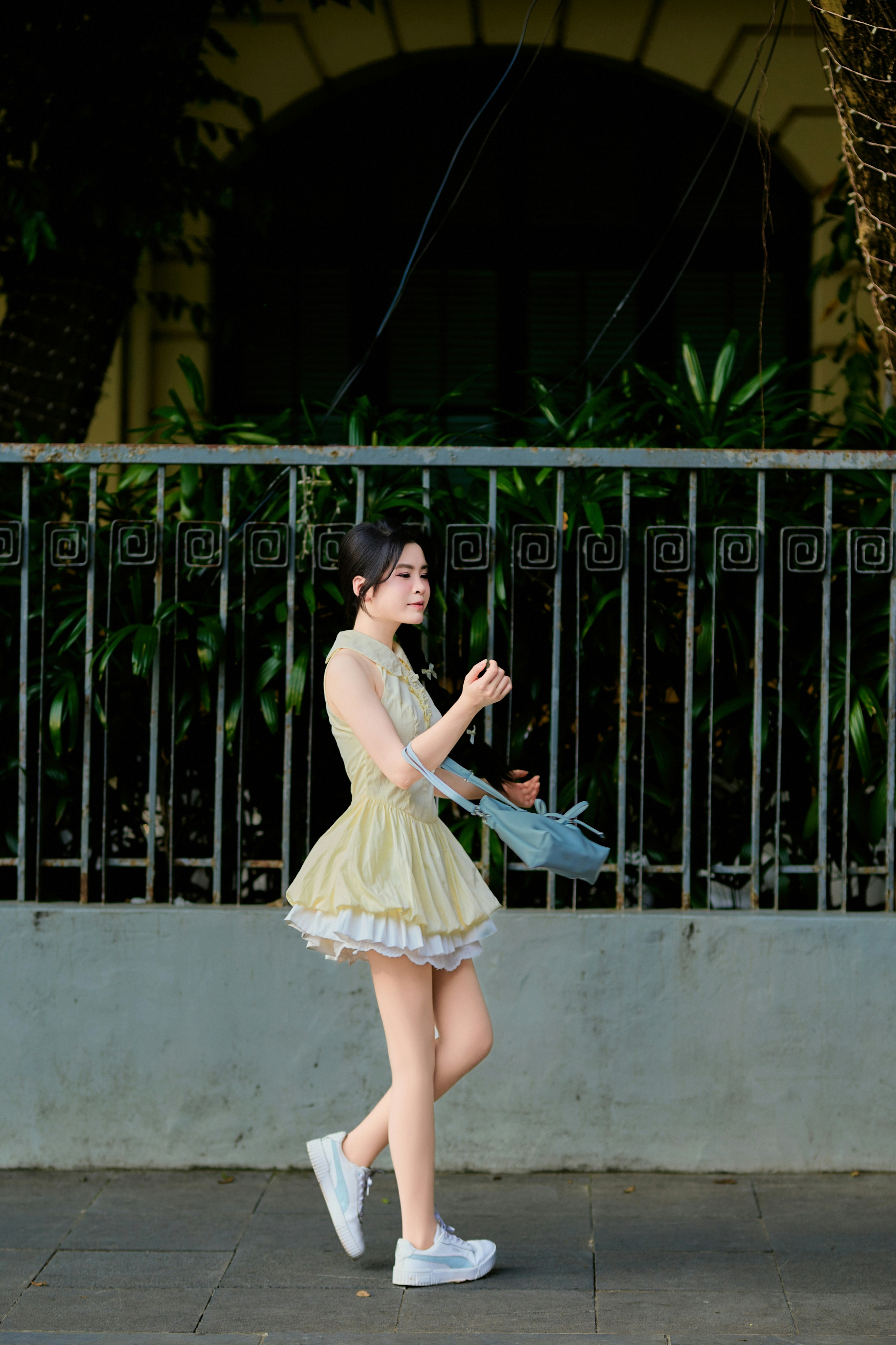 A young woman gracefully walking along a city sidewalk, dressed in a light-colored dress and holding an umbrella, framed by lush greenery and wrought iron railings.