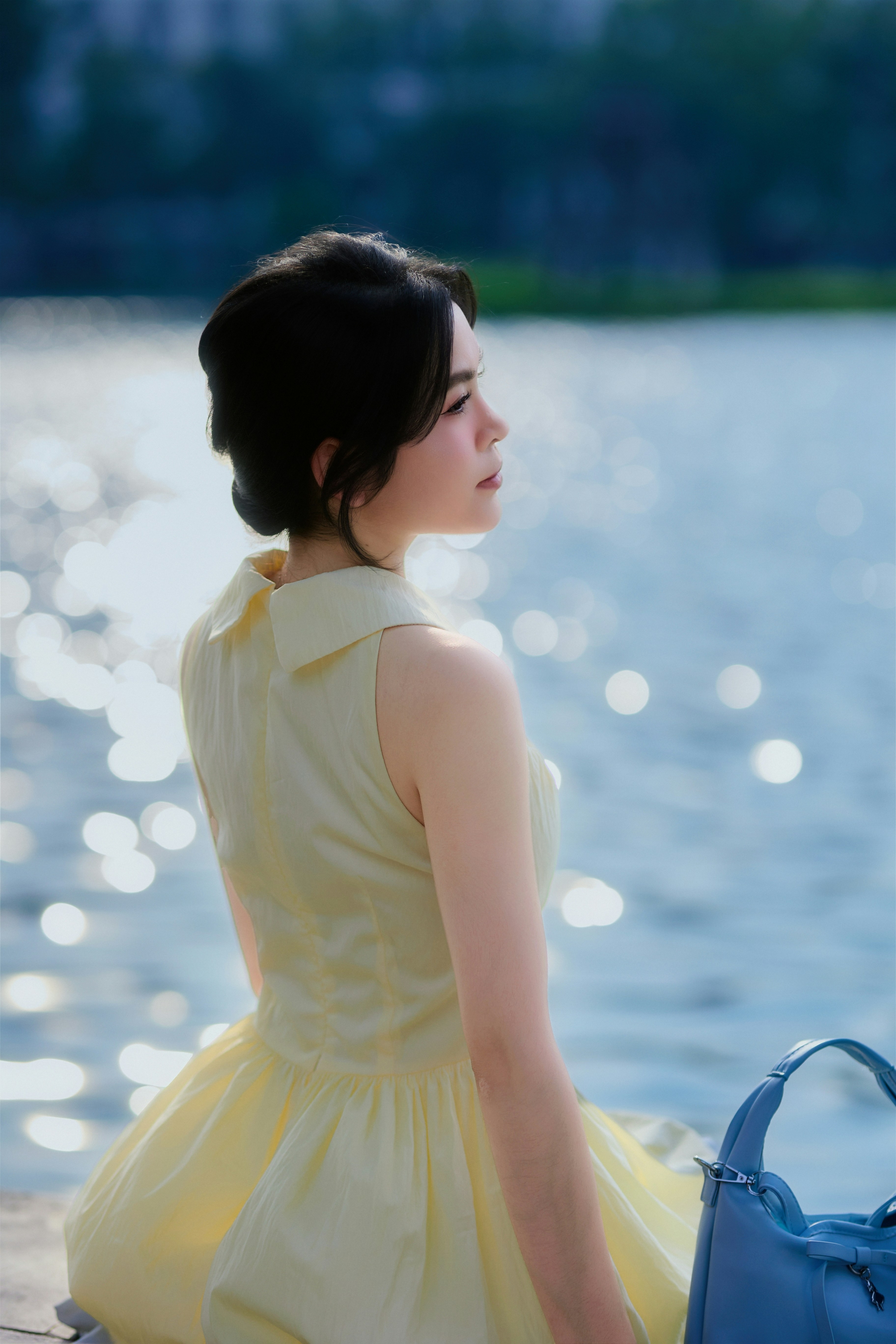 A woman in a yellow dress gazes thoughtfully at the shimmering water, with soft bokeh effects creating a tranquil atmosphere. Her blue handbag rests beside her.