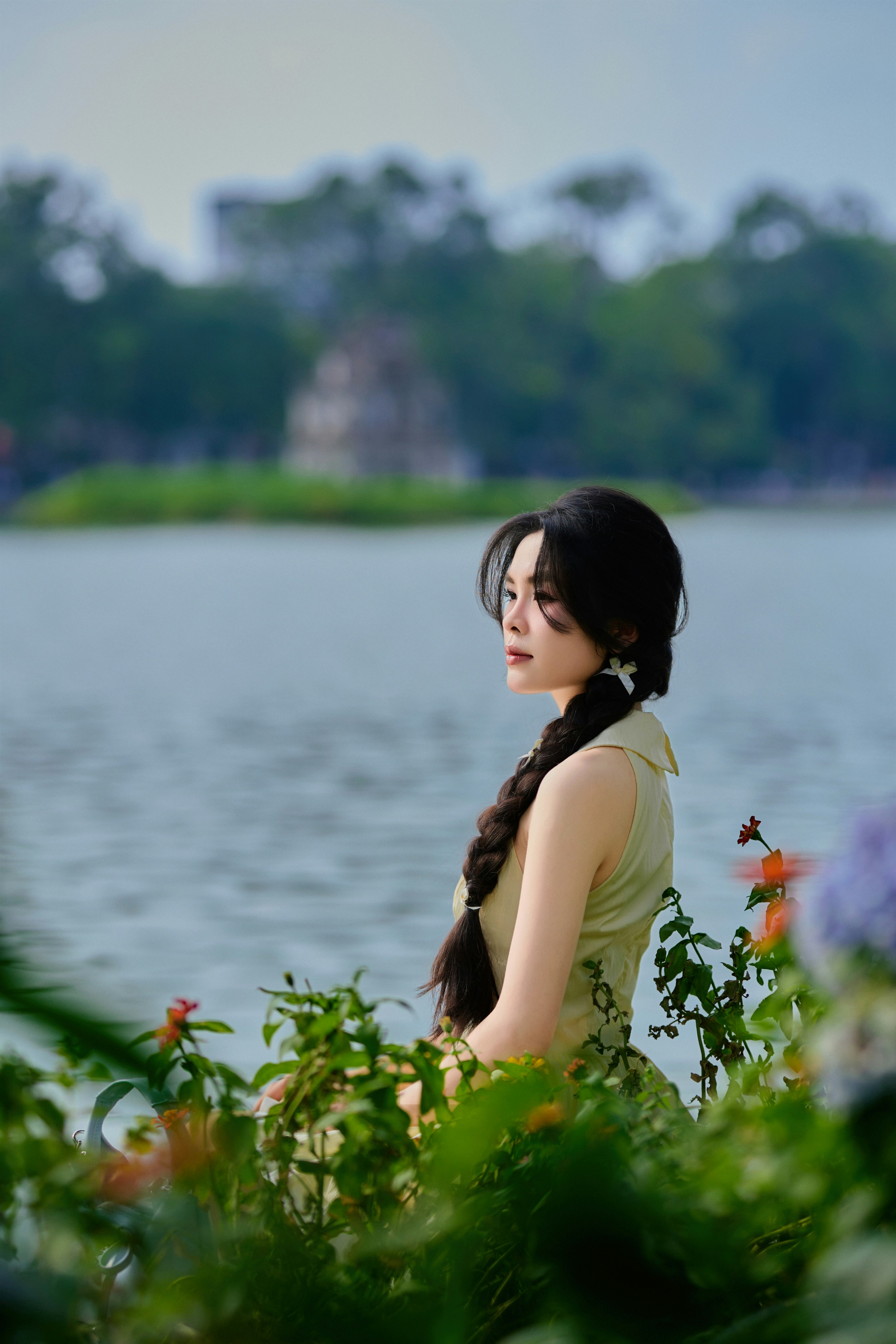 Young woman with a braided hairstyle sitting amidst vibrant flowers near a lake, gazing thoughtfully into the distance.
