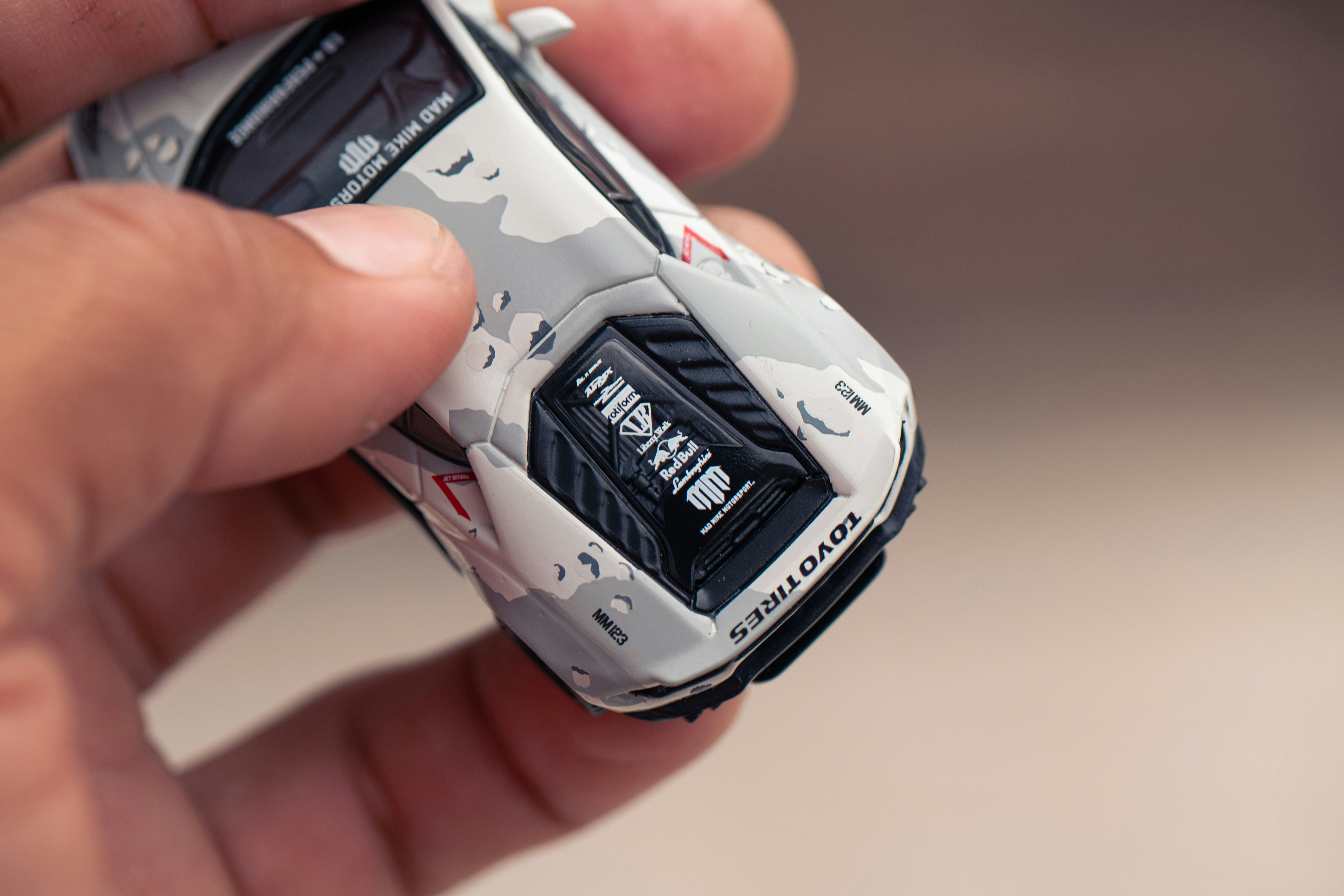 Hand holding a white toy car with camouflage pattern.