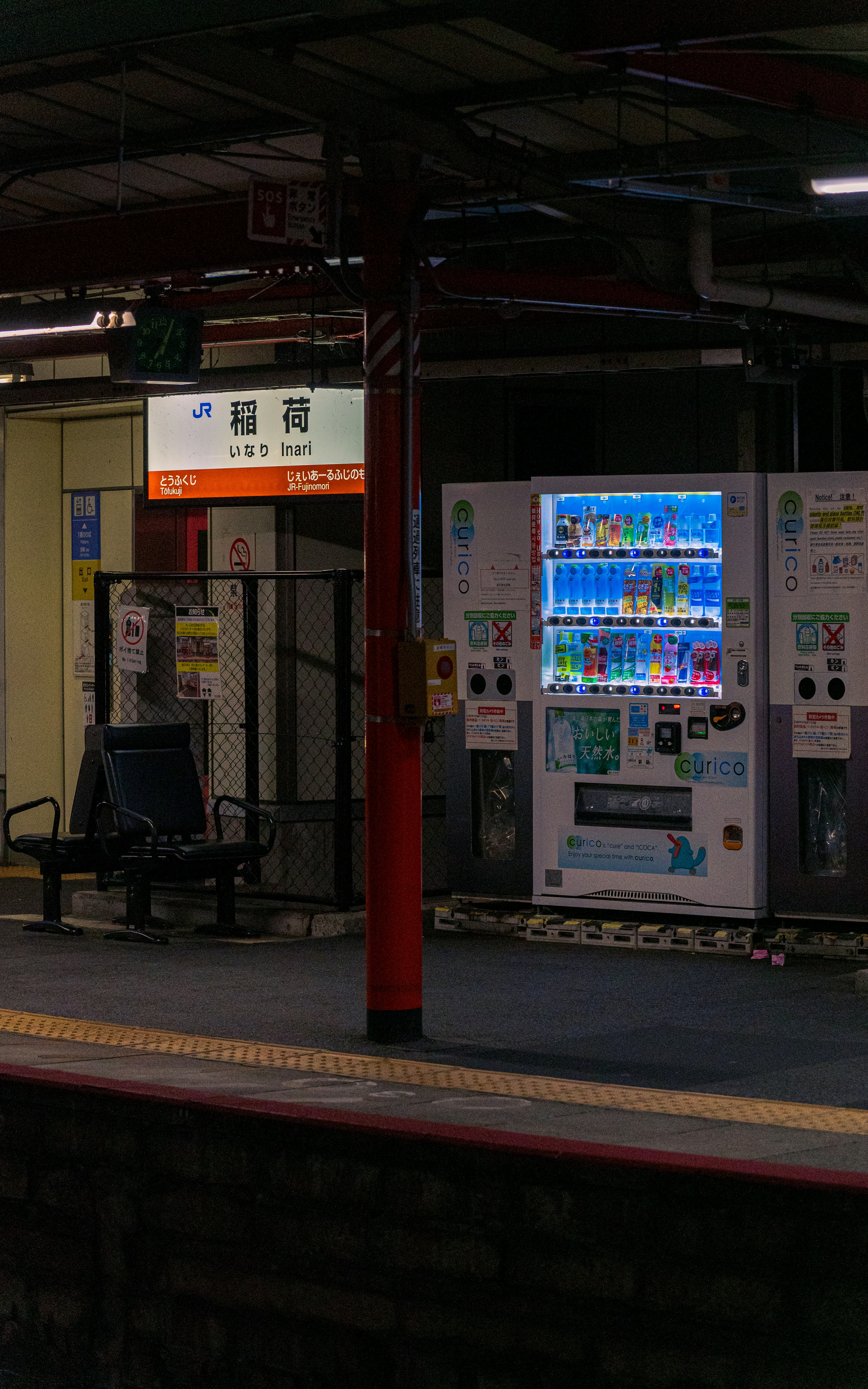 Vending machine glowing in the dimly lit Inari Station, surrounded by a quiet platform and signage. The scene captures the essence of late-night travel in Japan.