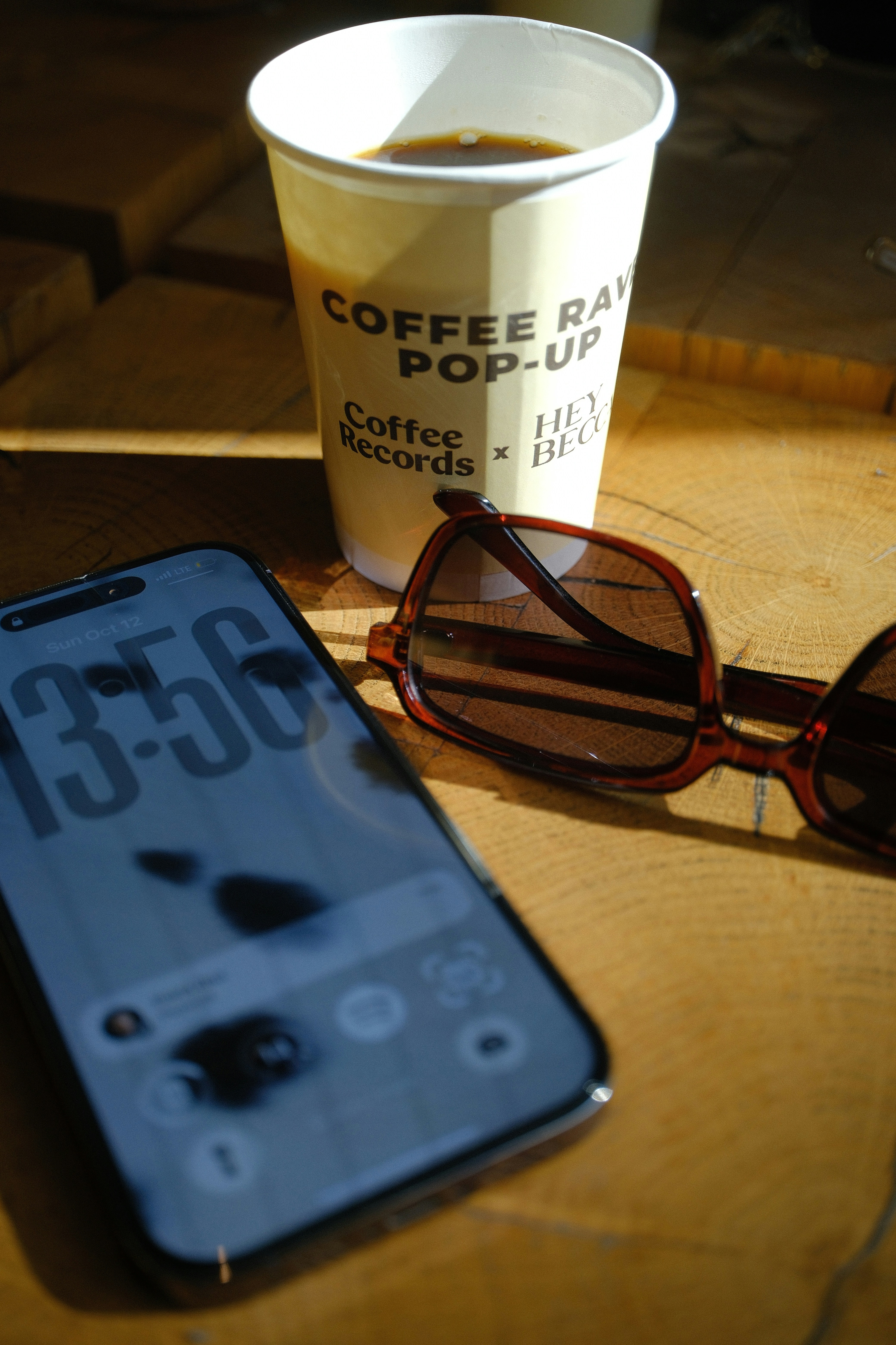 Smartphone, sunglasses, and coffee cup on wooden surface.