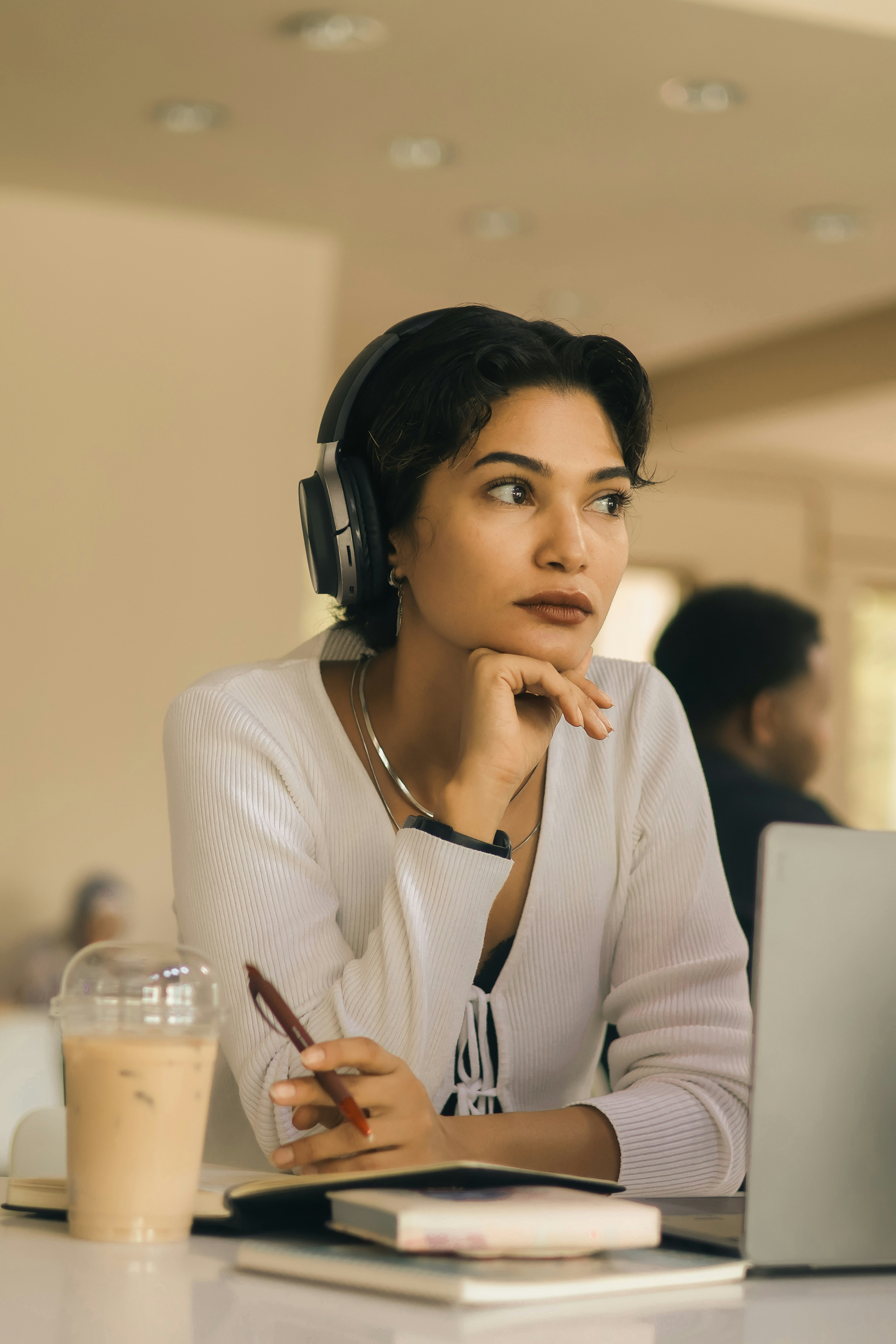 Young woman with headphones thoughtfully gazing while seated at a desk with a laptop and iced coffee. A notebook and pen are also visible.