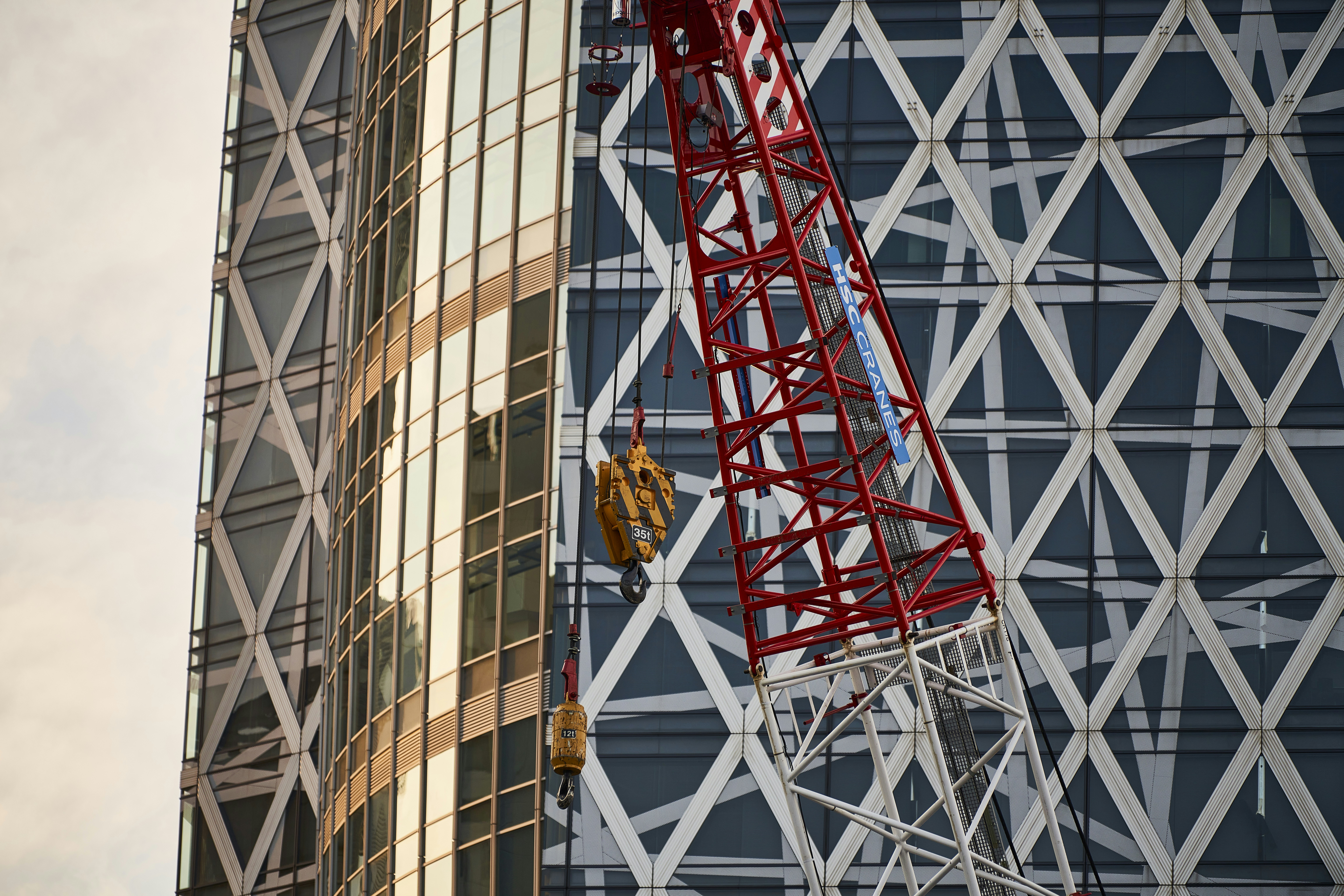A huge construction crane rests against the glass wall of a modern skyscraper nearing completion. The cool, geometrically patterned façade of the building contrasts sharply with the crane's rugged steel frame, painted a vibrant red. This is the dynamic moment when a city is growing and the landscape of the future is taking shape. This photograph is filled with the creative and developmental energy inherent in the act of construction. | Construction crane against modern glass building