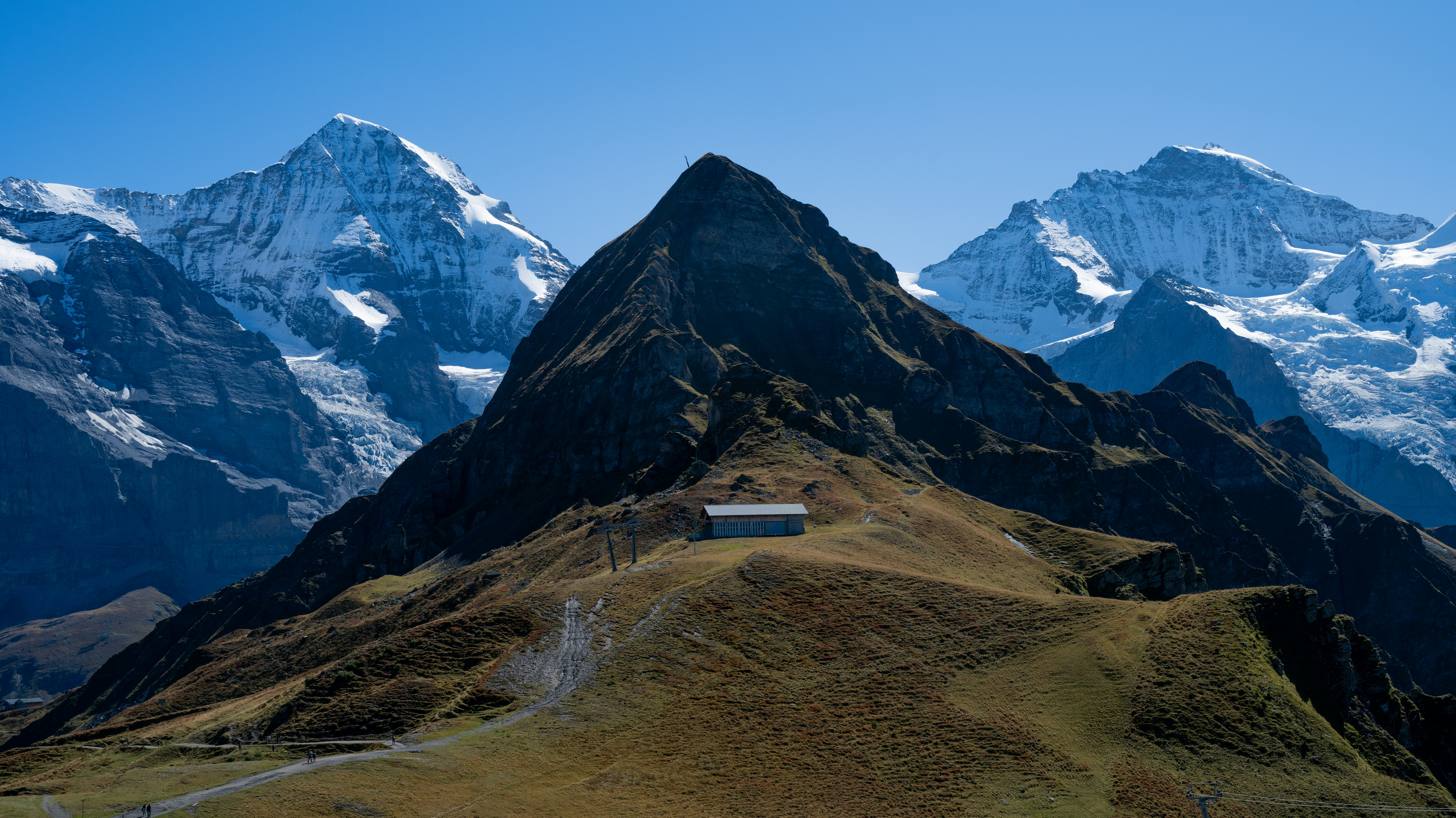 A solitary structure rests on a grassy knoll amidst towering snow-capped peaks, showcasing the stark contrast between man-made and natural beauty.
