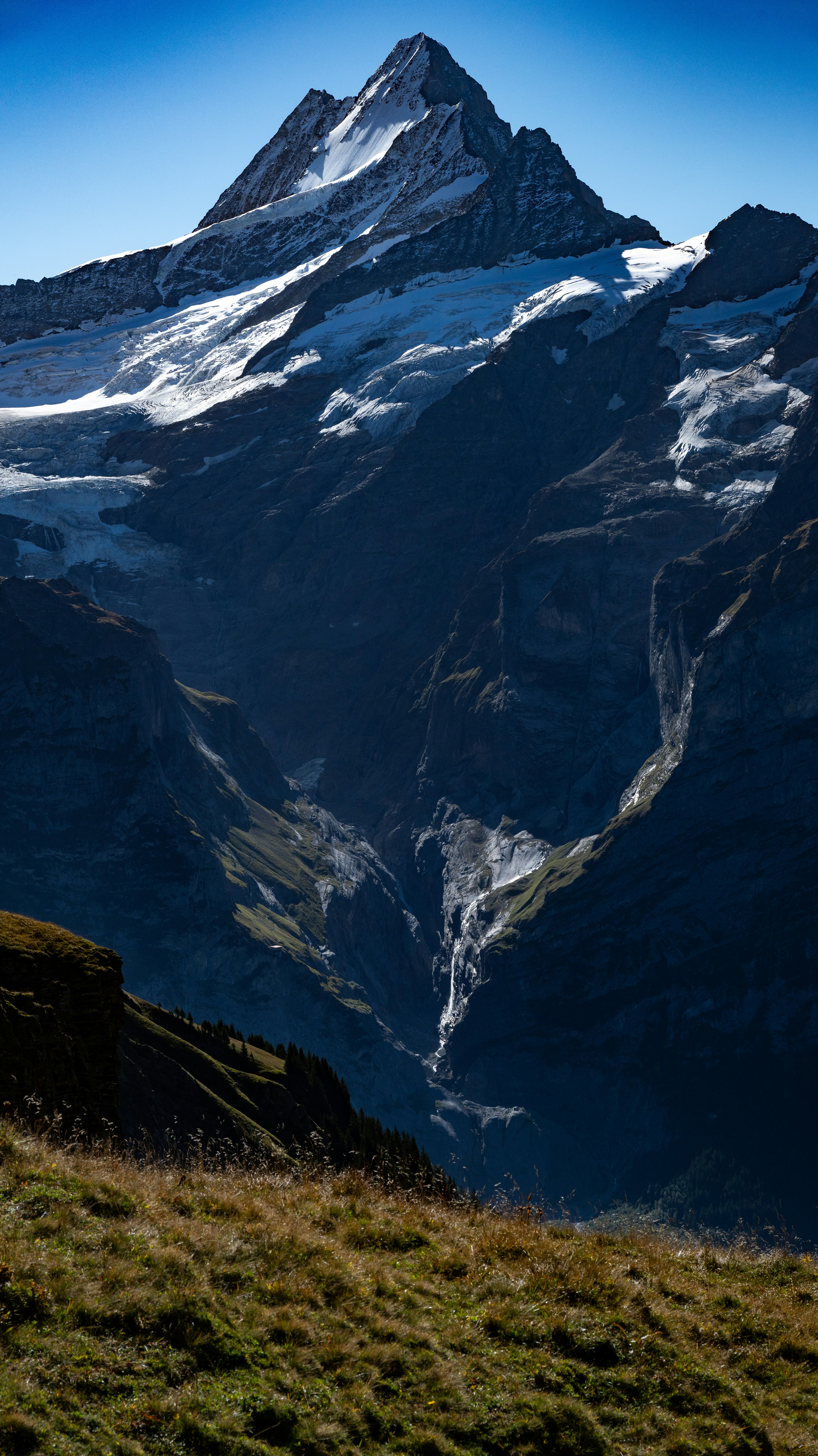 A snow-capped mountain peak towers above a deep valley, with a cascading waterfall visible in the shadows below. The rugged terrain showcases the raw beauty of nature.