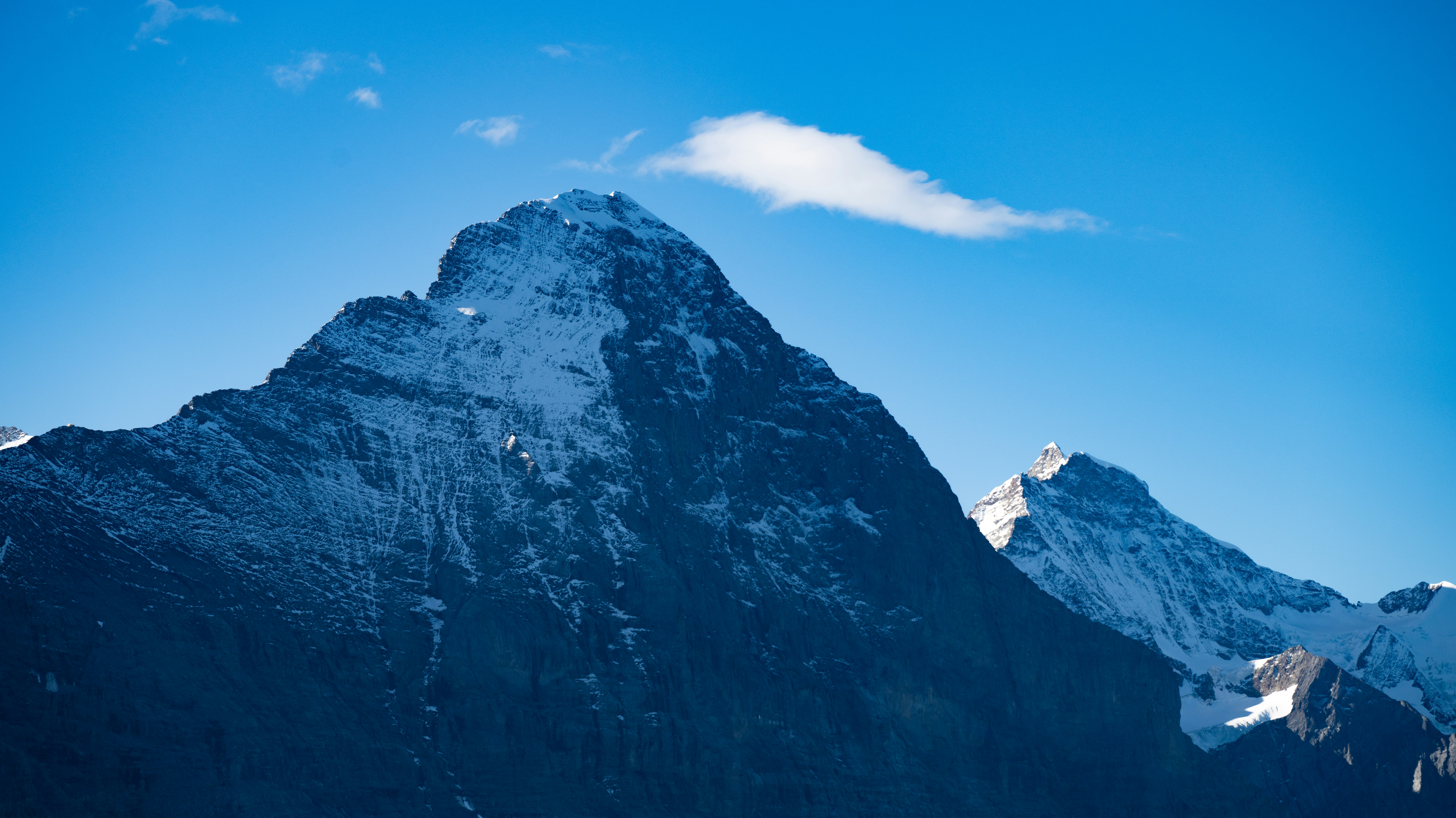Snow-capped mountain range under a bright blue sky, showcasing the rugged beauty of nature. The sunlight highlights the texture of the rocky surfaces.