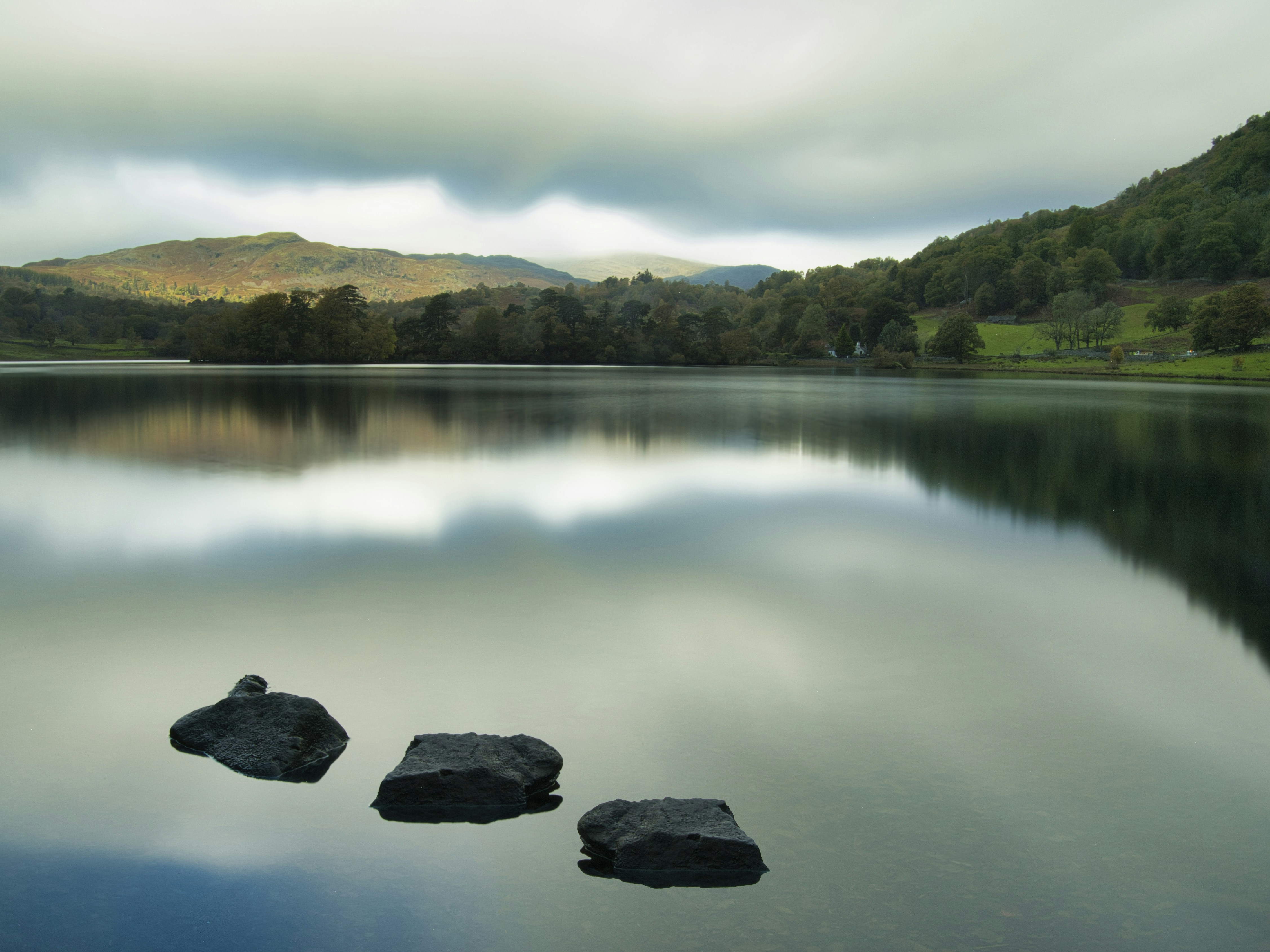 Three smooth stones partially submerged in a serene lake, reflecting the soft hues of the cloudy sky and surrounding hills.
