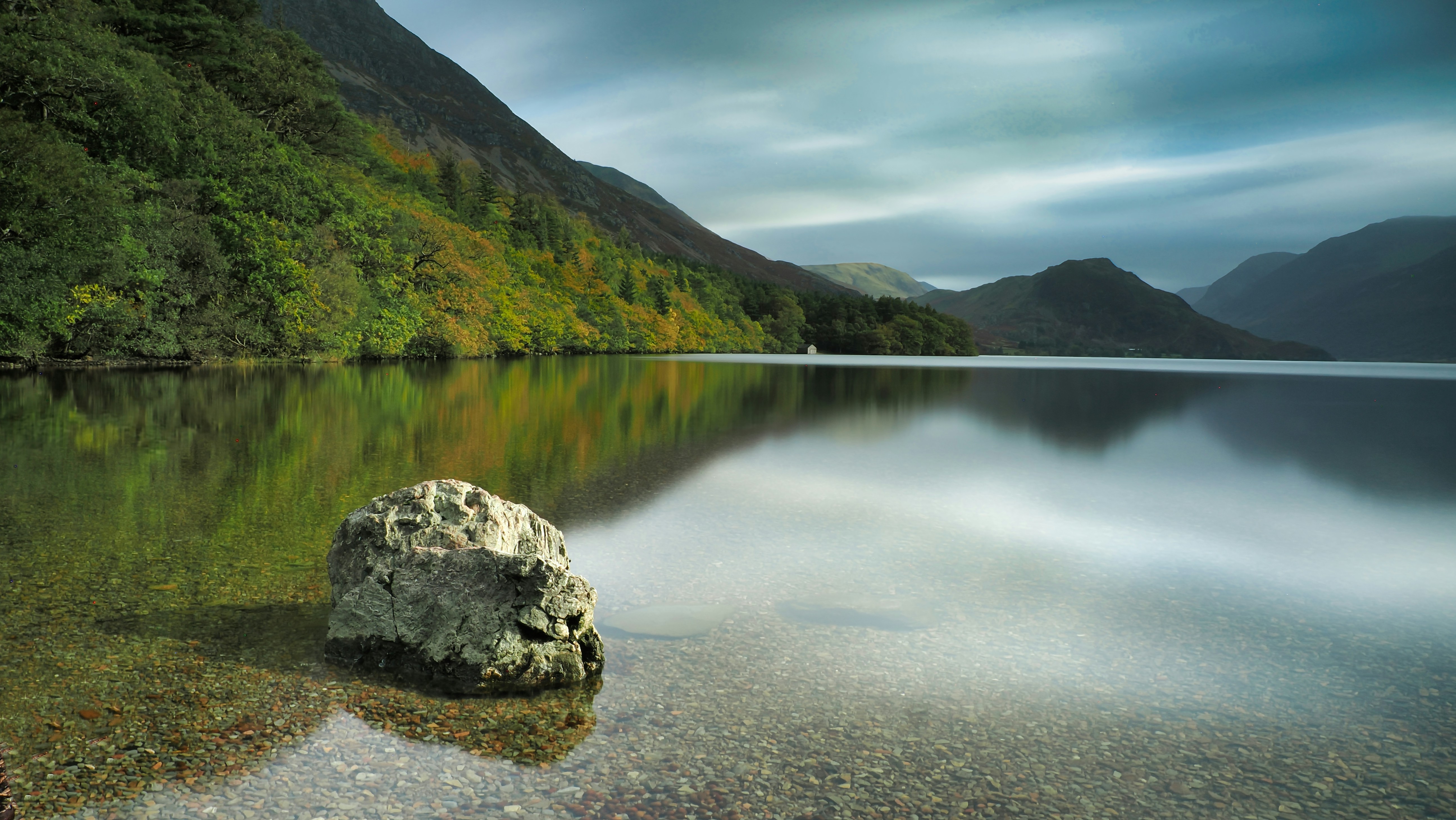 Smooth lake surface reflecting surrounding hills and a solitary rock in the foreground. The tranquil scene invites contemplation.