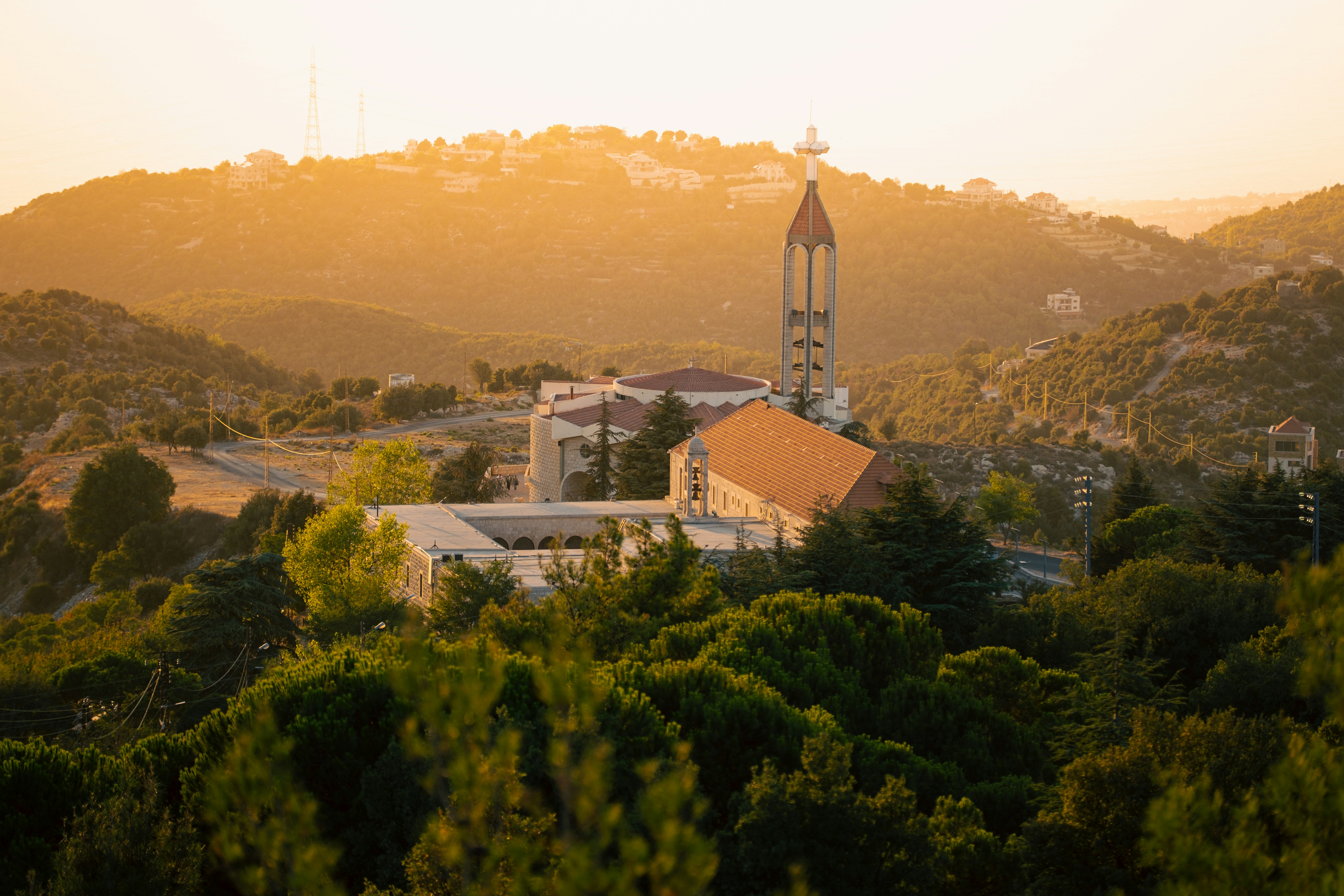 A hillside church bathed in warm golden light during sunset, surrounded by lush greenery and distant hills. The scene captures the tranquility of nature and architecture harmoniously coexisting.