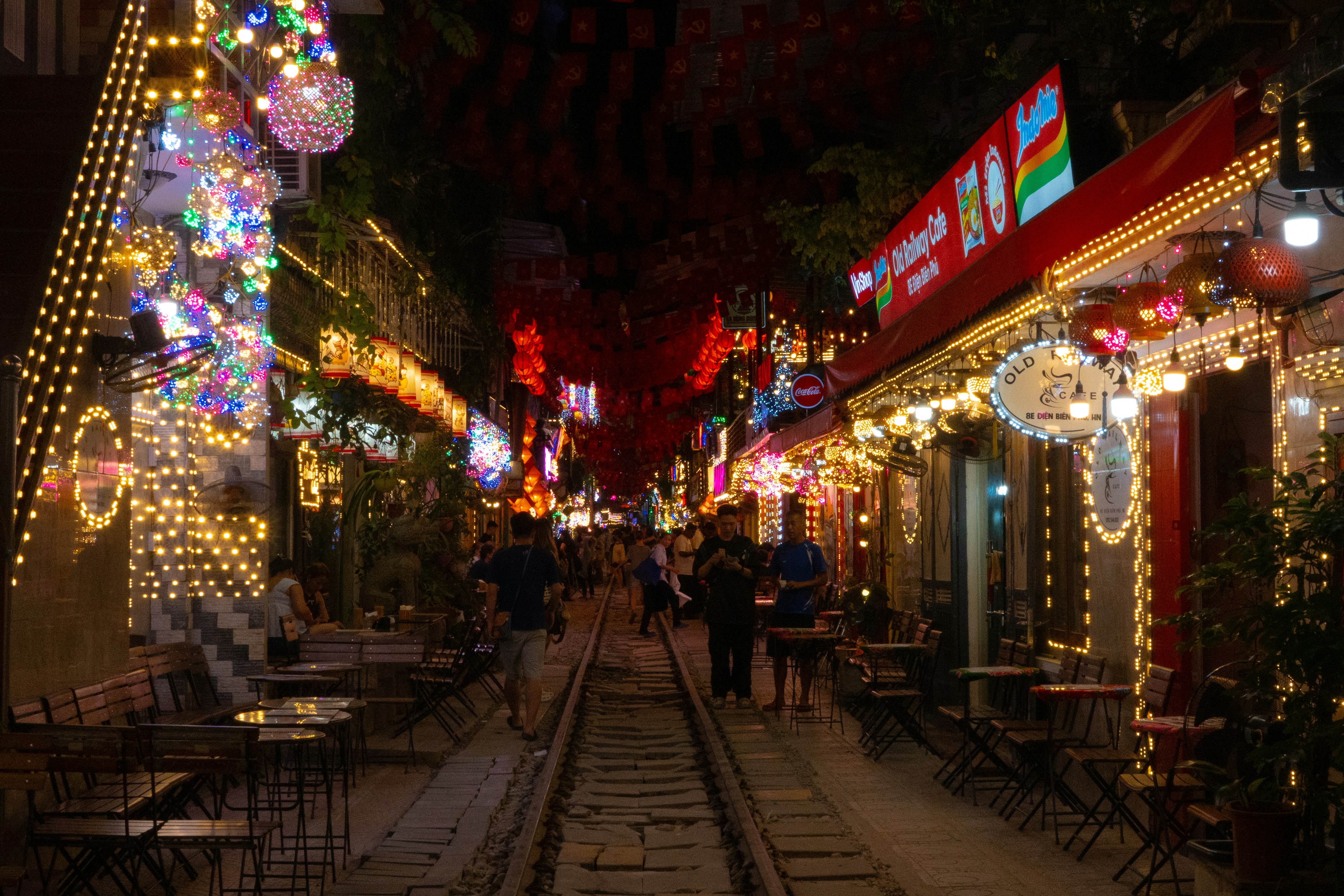 A vibrant street lined with colorful lights and bustling cafes, featuring railway tracks that add a unique charm to the scene.