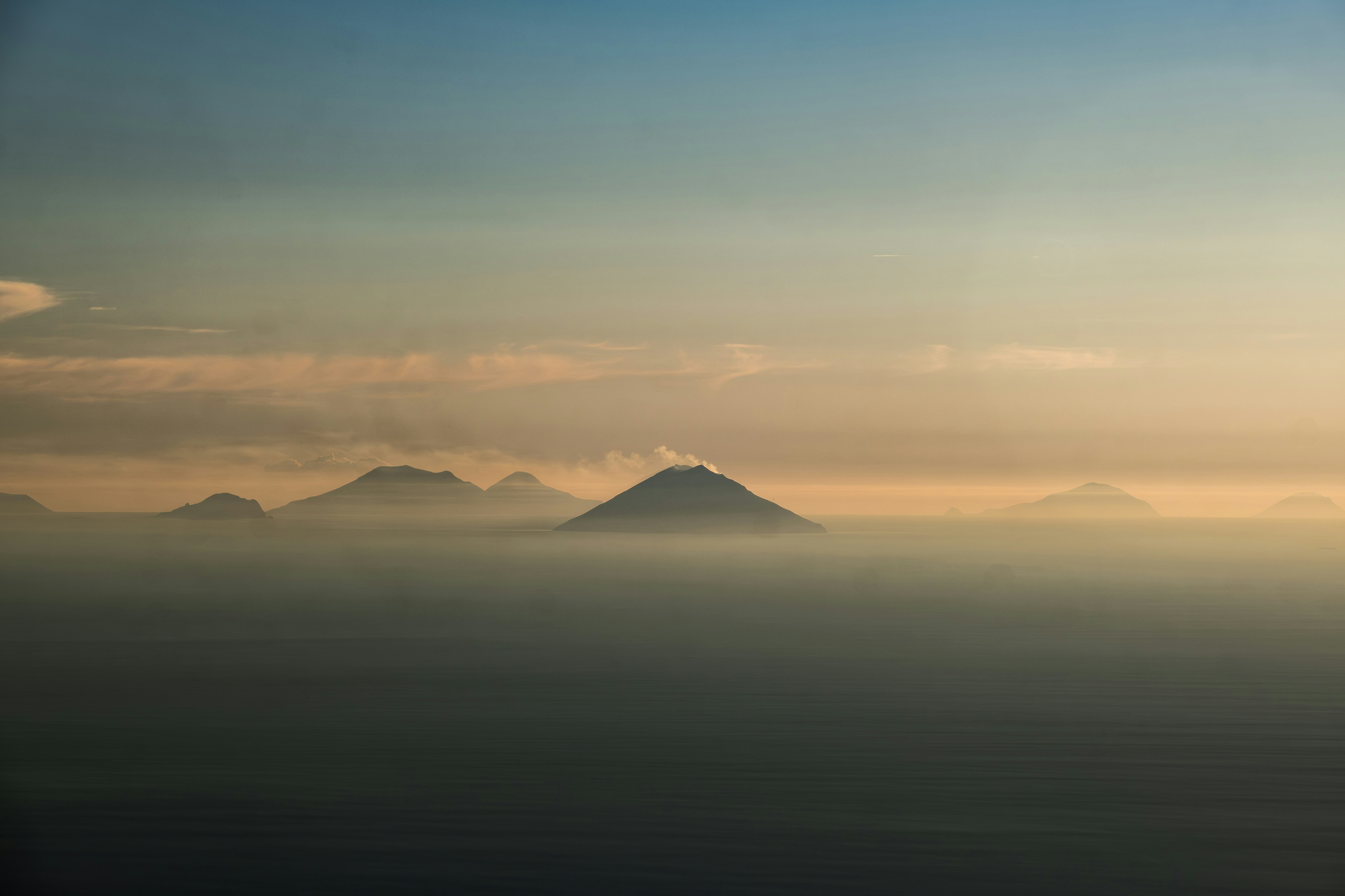 A breathtaking view of volcanic islands at sunset, emerging through a soft mist over the sea. The central volcano gently releases smoke into the pastel-colored sky, creating a serene and otherworldly atmosphere. | Islands emerging from mist at sunrise